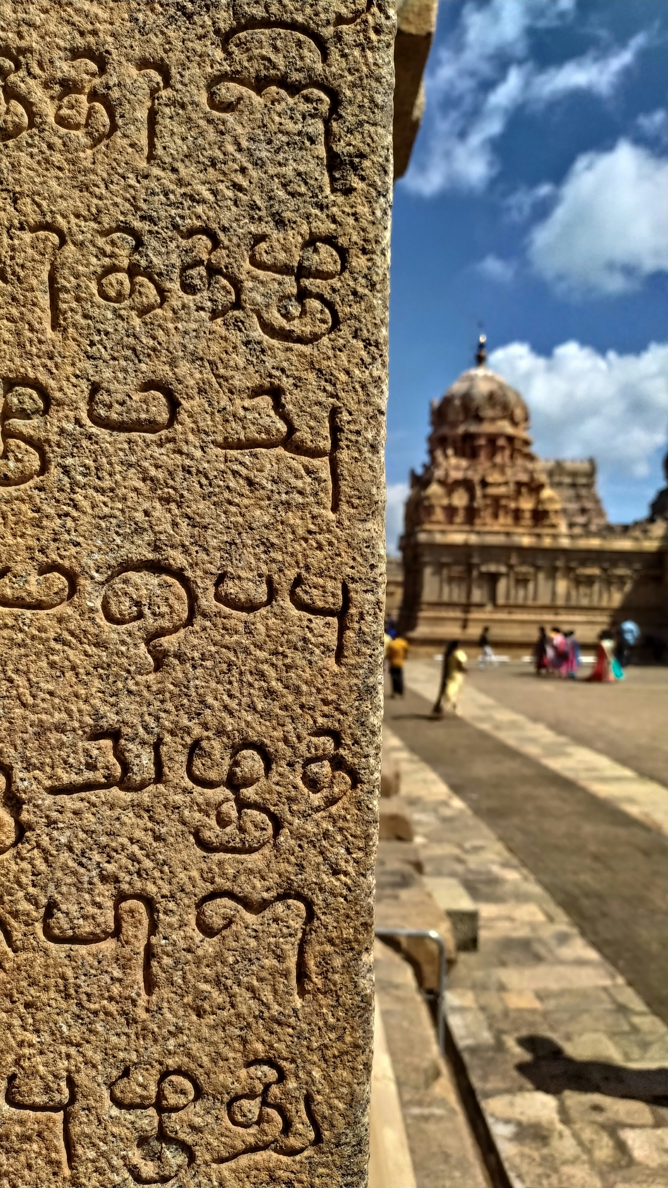 Close-up of a weathered stone pillar with carved script, leading the viewer's eye toward a distant temple and visitors in a sunlit courtyard.