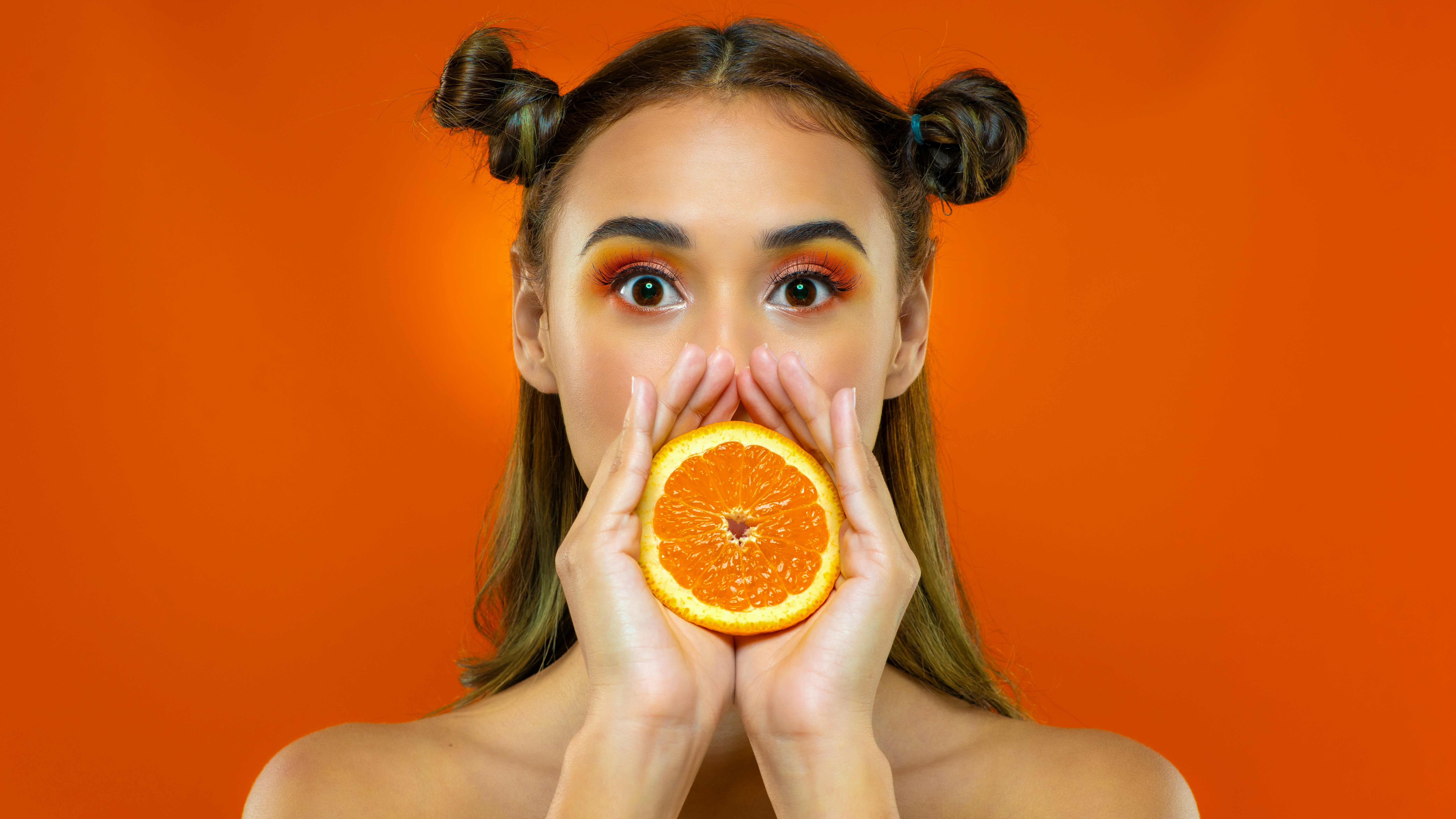 Woman with orange slice against a vivid orange background.