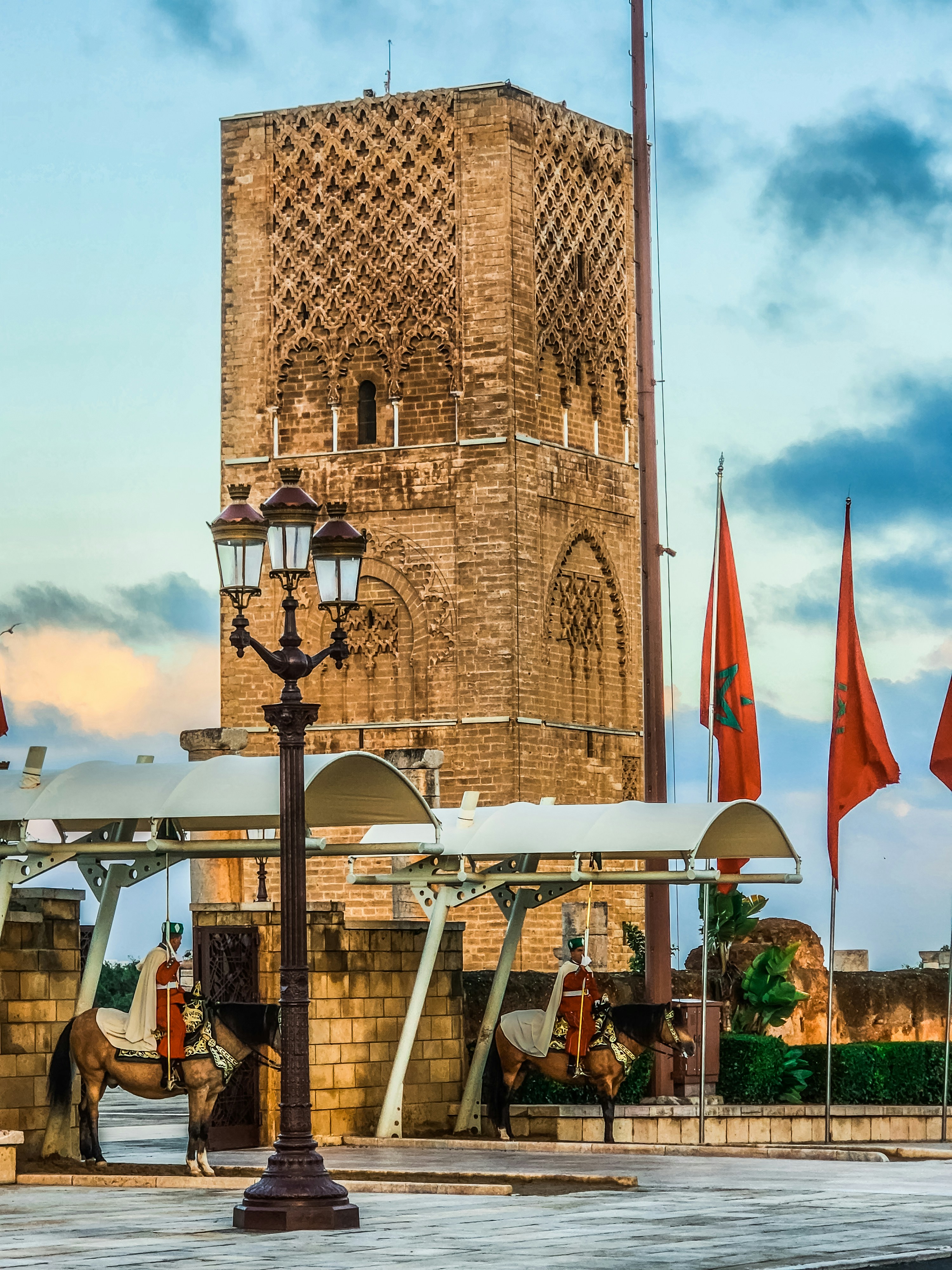 Historic tower adorned with intricate designs, flanked by regal horses and Moroccan flags. A street lamp adds charm to the scene.