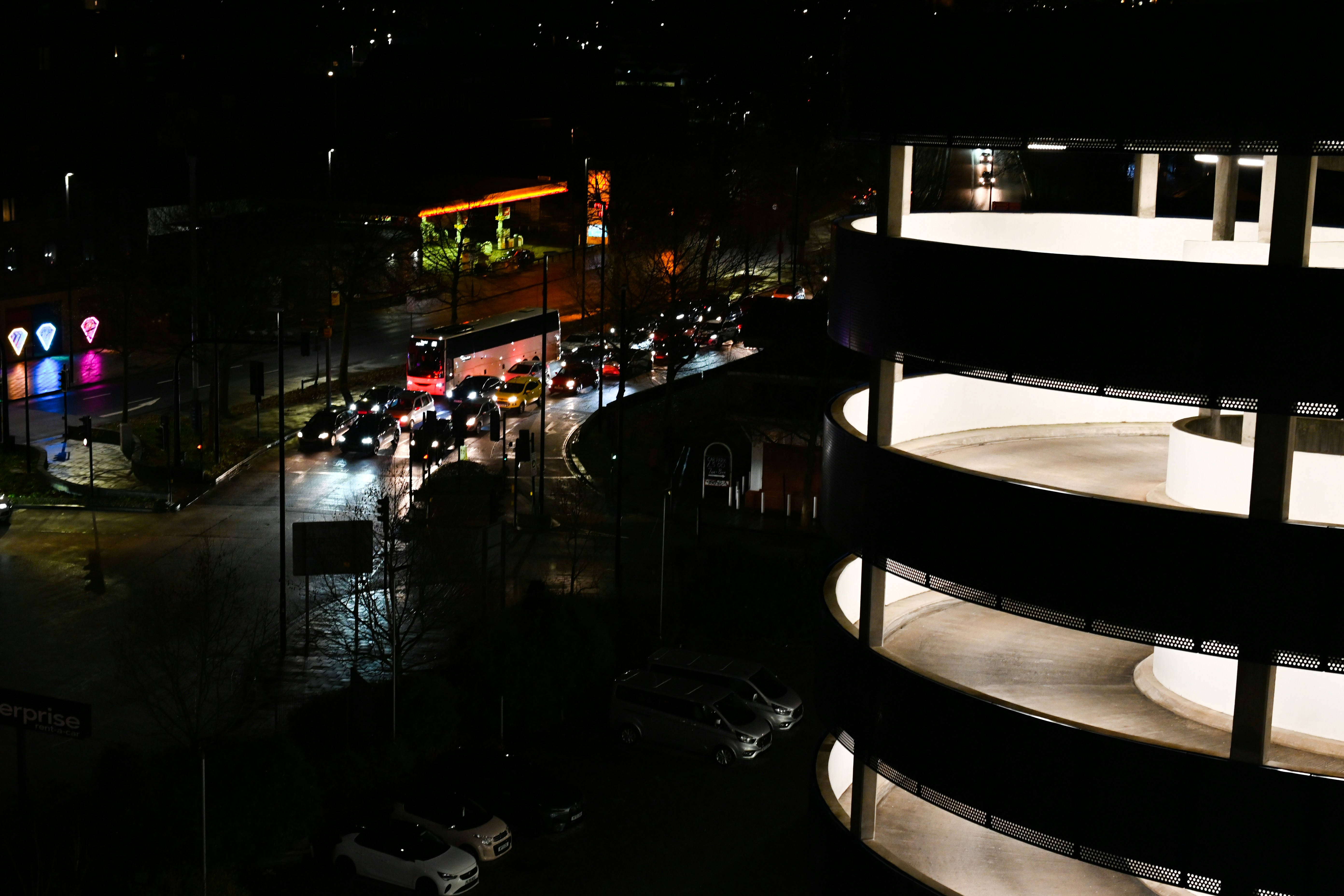 A vibrant city scene at night featuring illuminated traffic and a modern parking structure. Colorful lights reflect off the wet pavement.