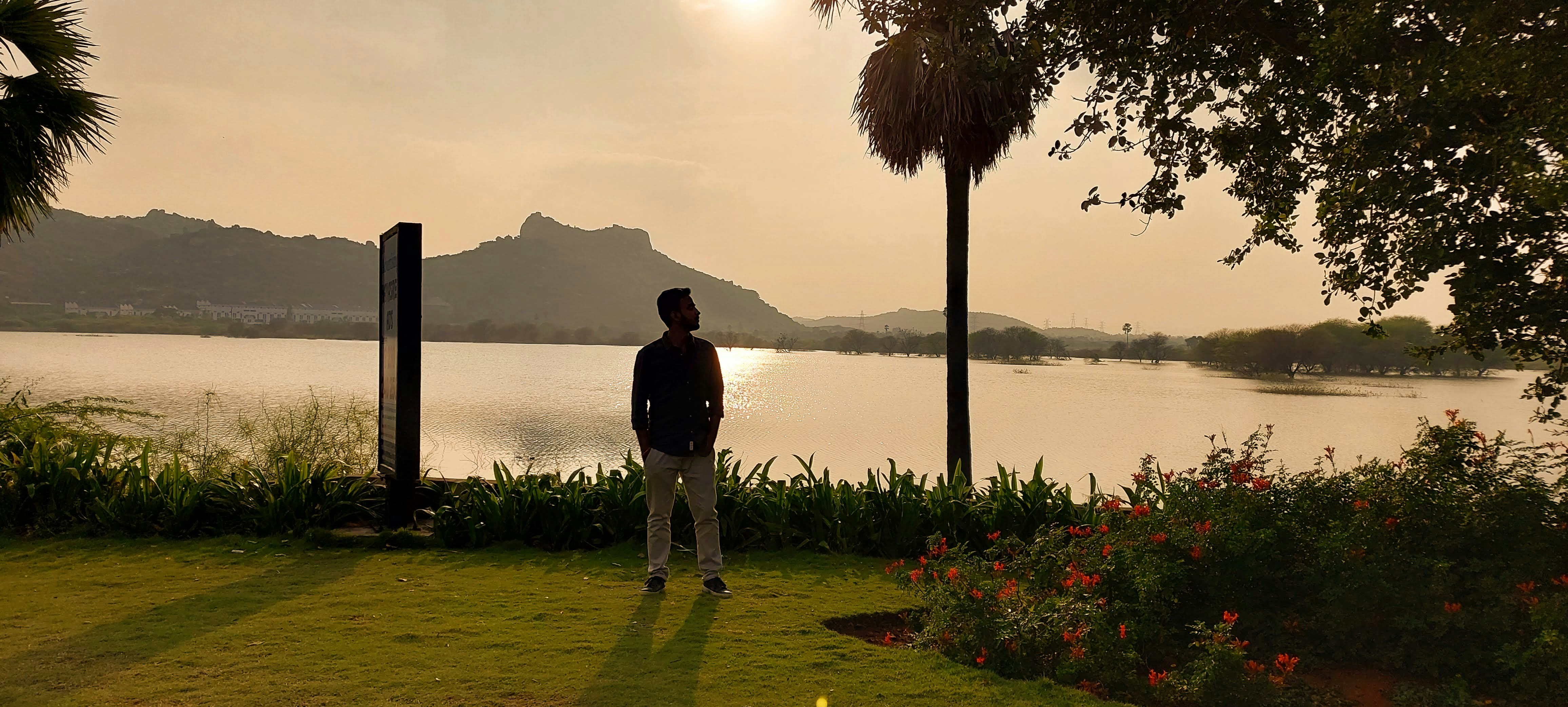 Lone figure stands on a grassy bank beside a calm lake at sunset, with distant hills and a tree-framed shoreline.