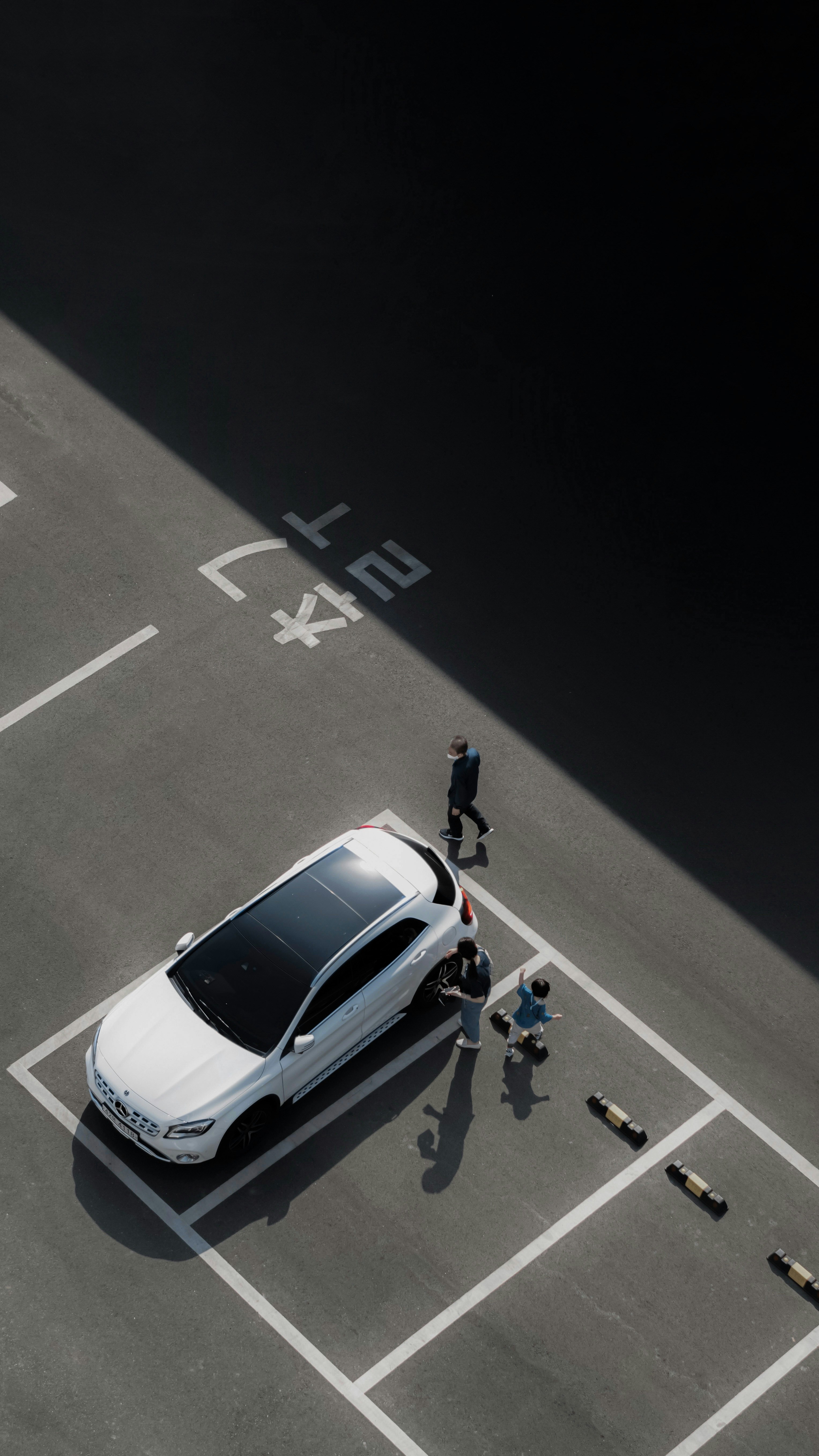A family interacts near a white SUV in a parking lot, with shadows creating a striking contrast against the pavement.