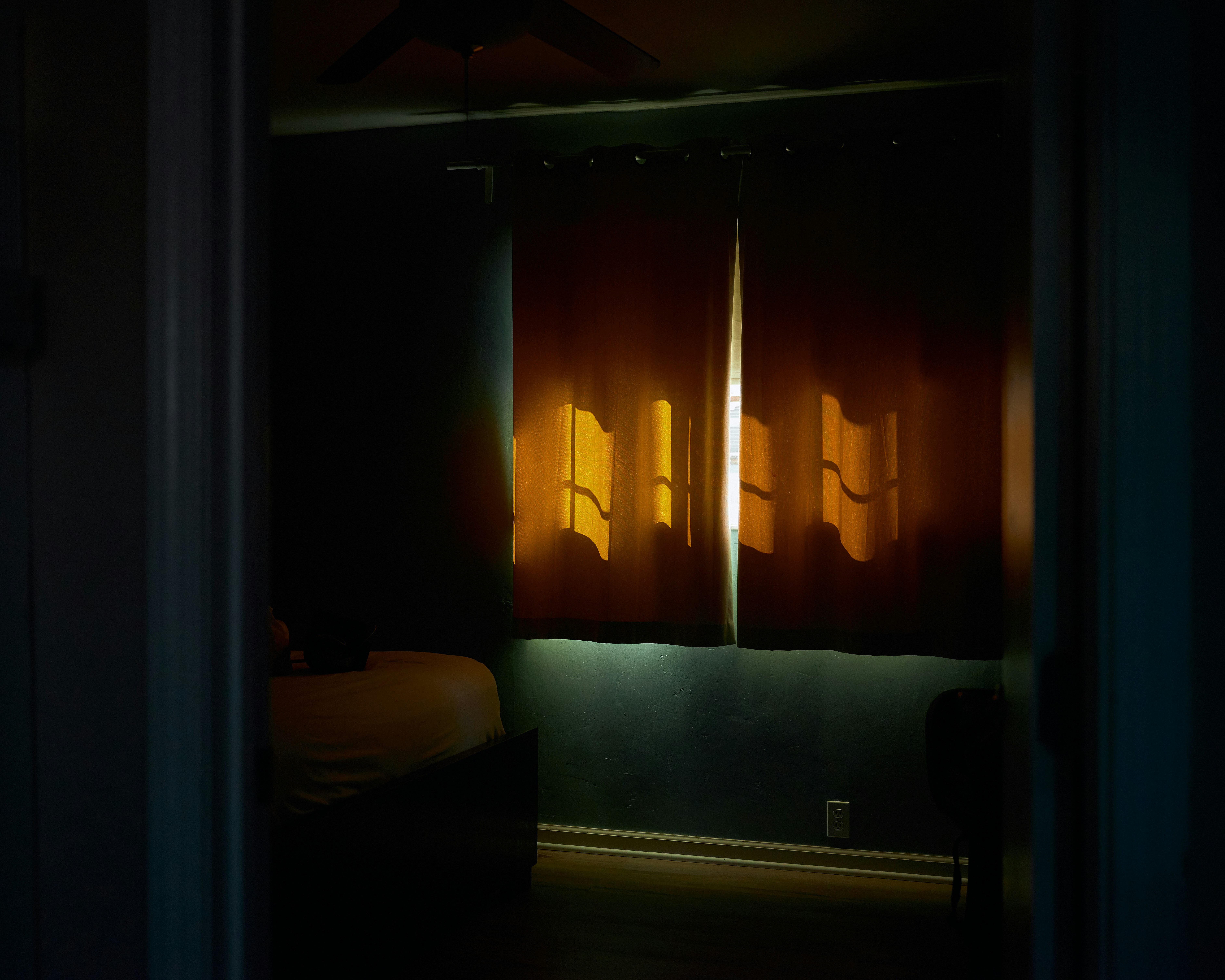 Soft morning light filters through dark curtains, casting intricate patterns on the wall in a serene bedroom setting.