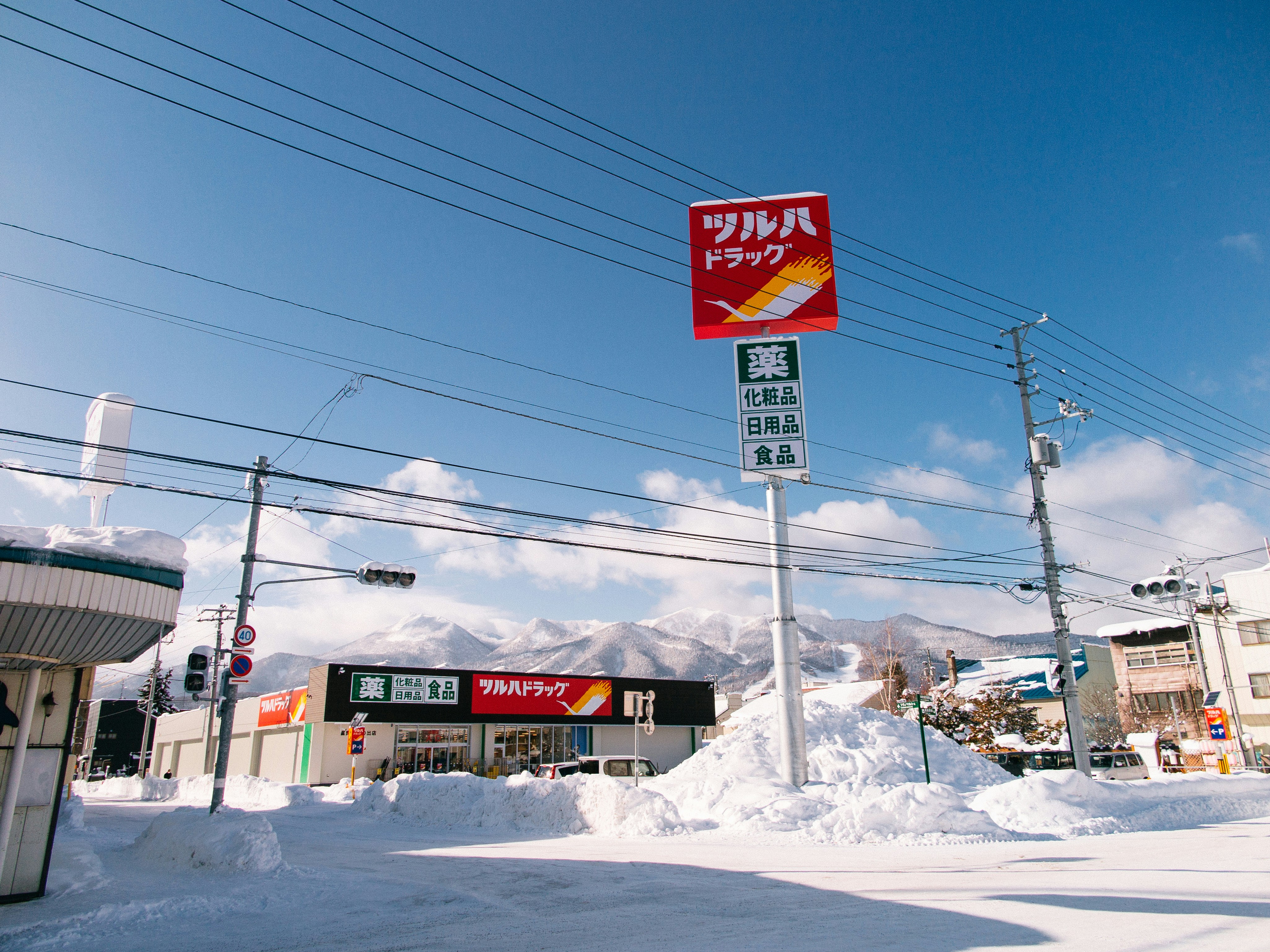 Snow-covered street with a tall red sign towering over storefronts, set against a bright blue sky and distant mountains.