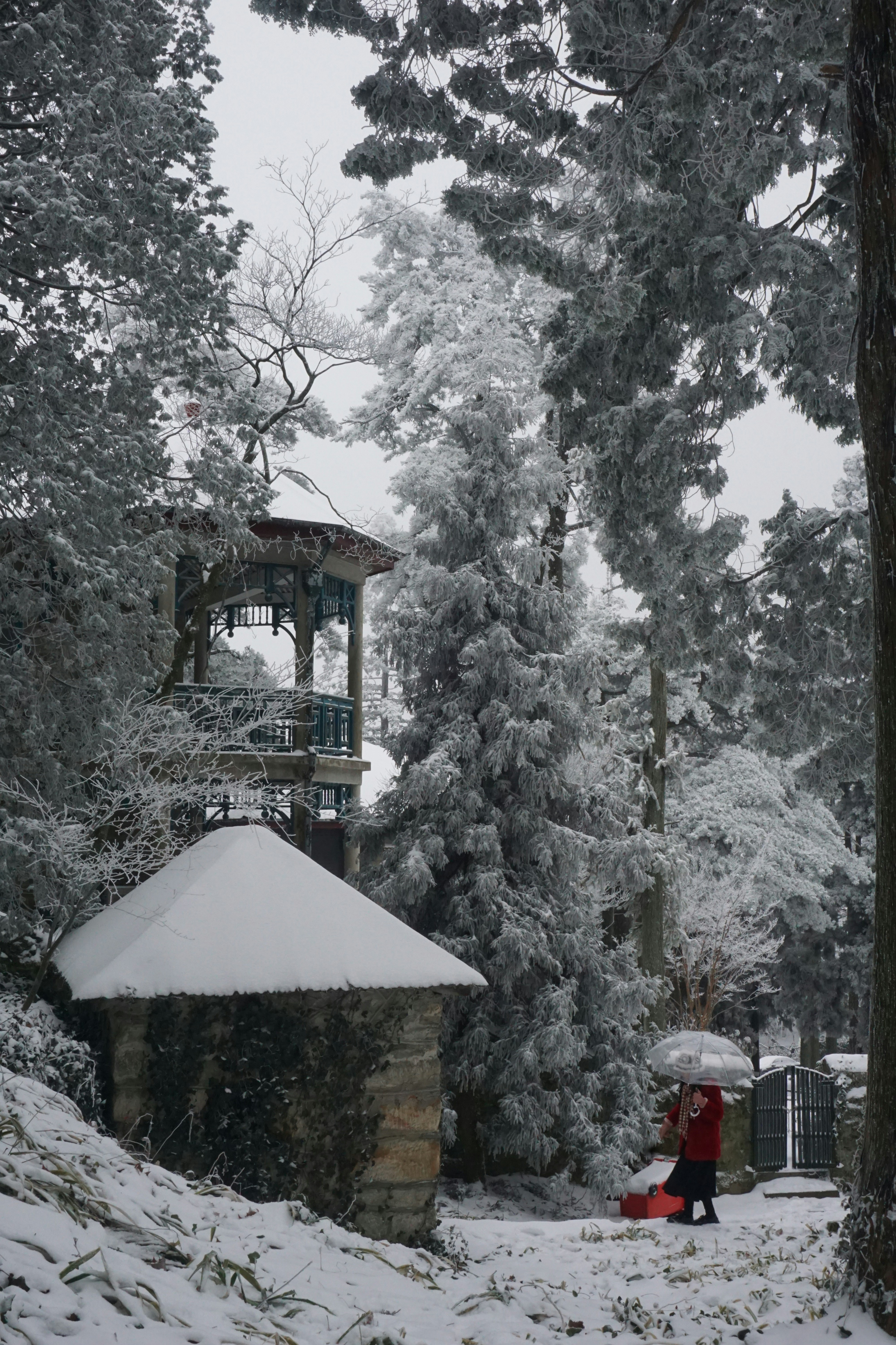 Person in red coat and umbrella walking through a snow-laden forest path beside a stone structure and wooden gazebo.