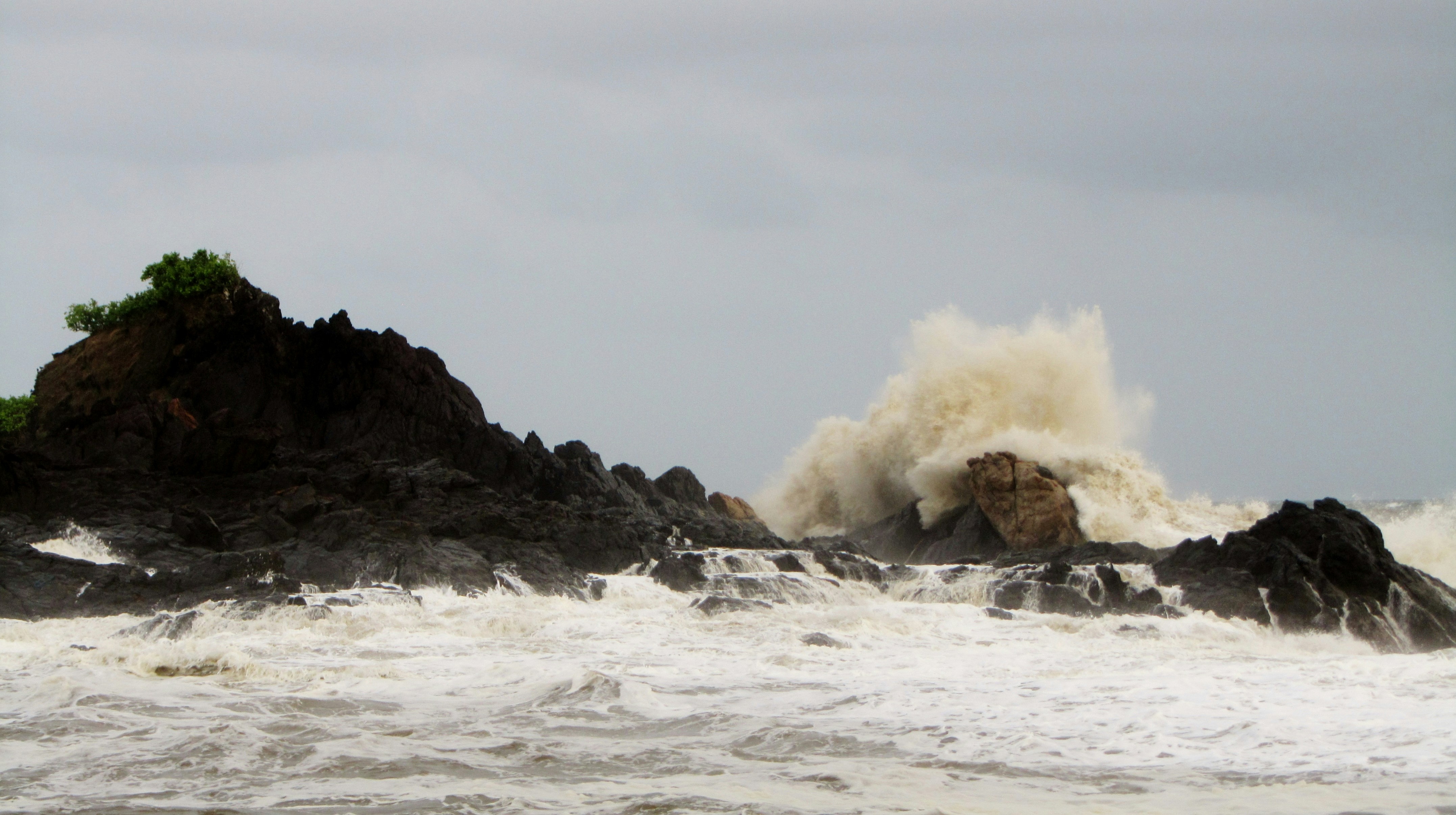 Powerful waves crash against jagged black rocks near a windswept shore, sending spray into the air. A small green outcrop anchors the left side of the scene.