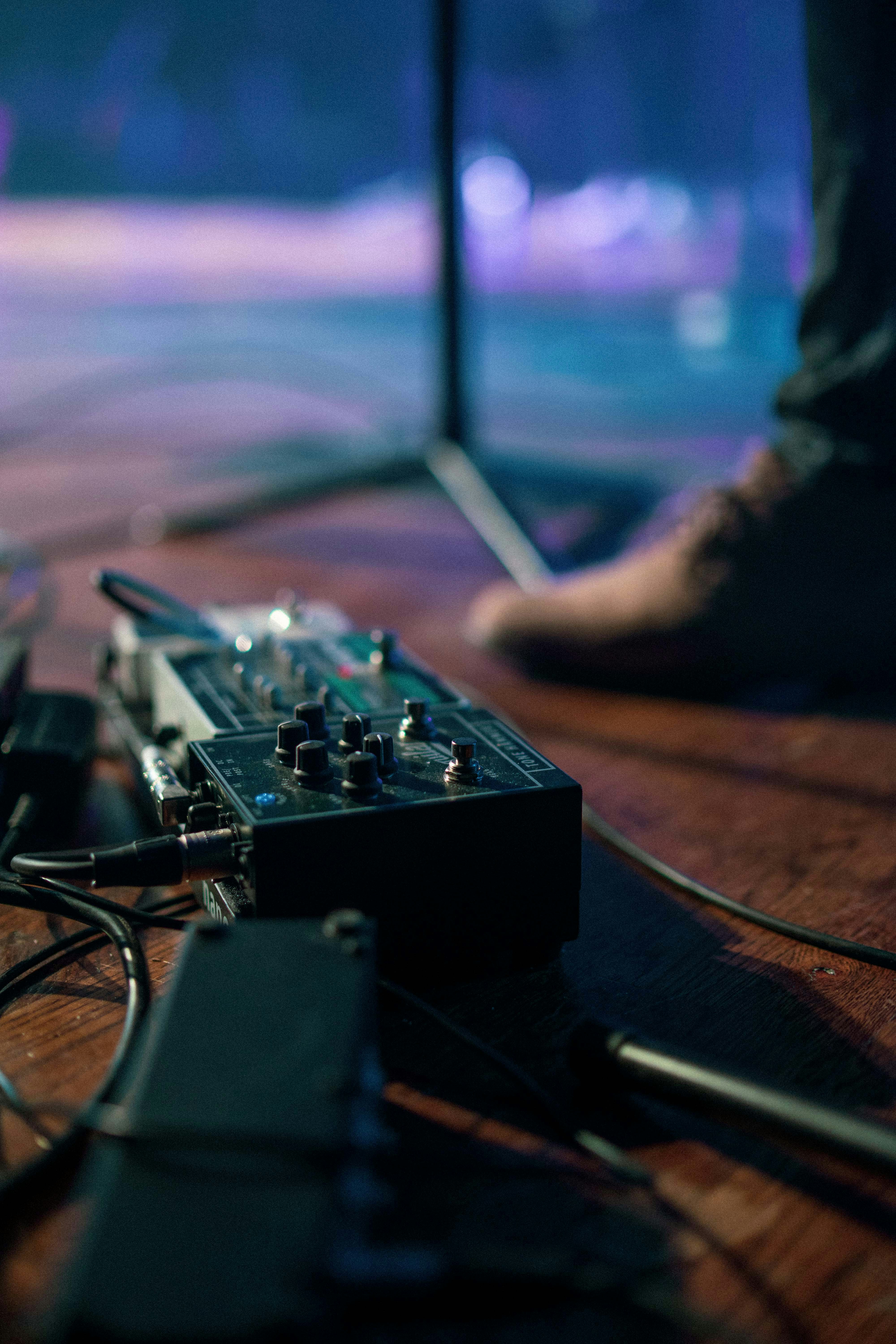 Close-up of a guitar effects pedal on a wooden stage, with a musician's foot poised nearby. The vibrant lighting creates an atmospheric backdrop.
