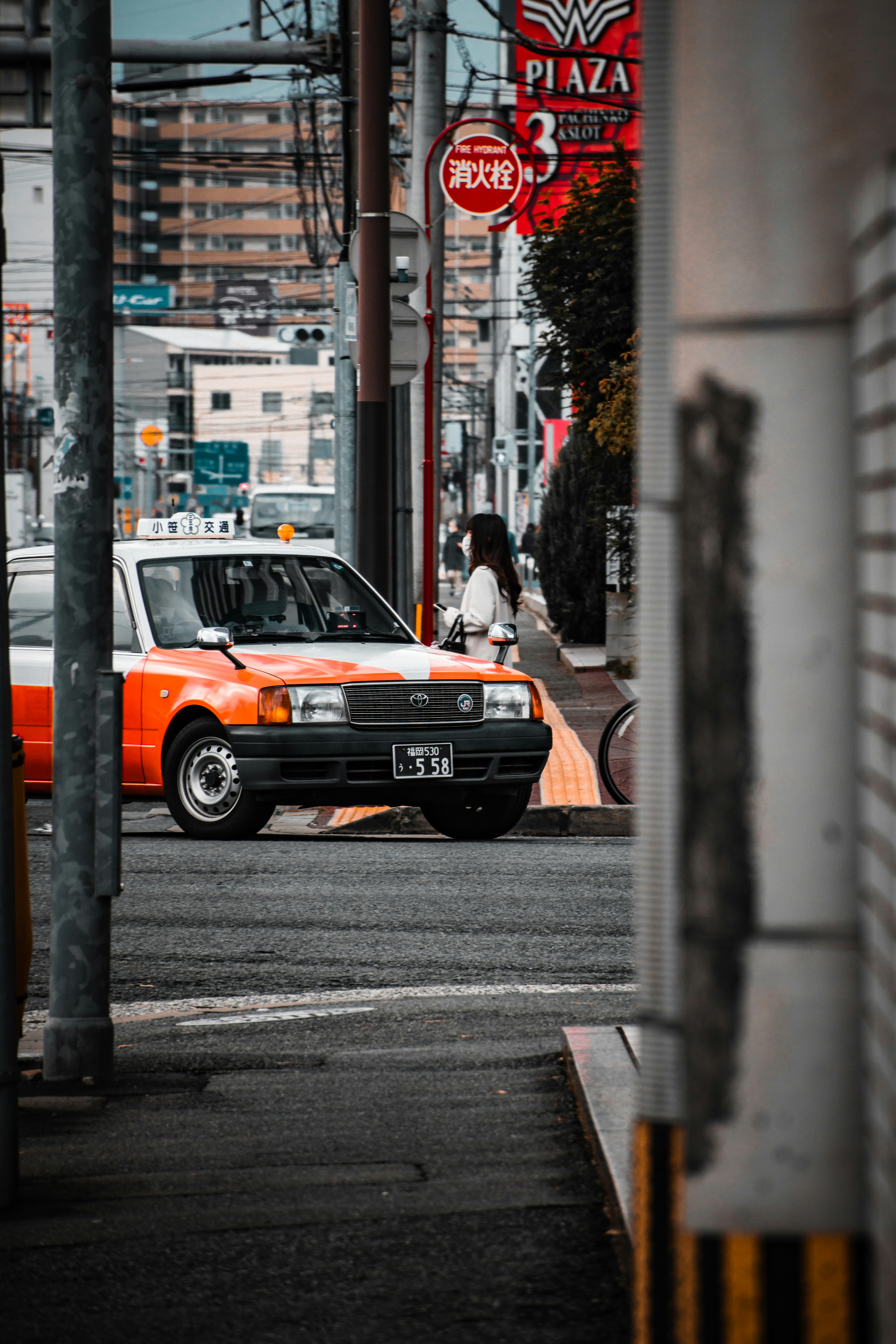 Orange taxi navigating a bustling street corner, framed by urban elements and a passerby. The scene captures the essence of city dynamics.