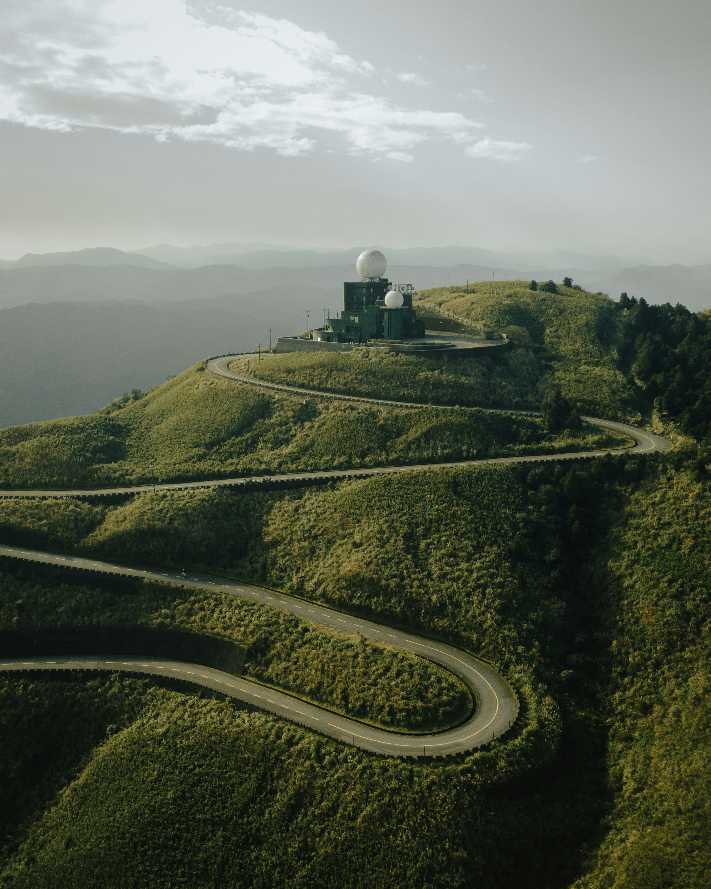 Aerial view of a winding road leading to a radar station atop a green hill, surrounded by misty mountains in the background.