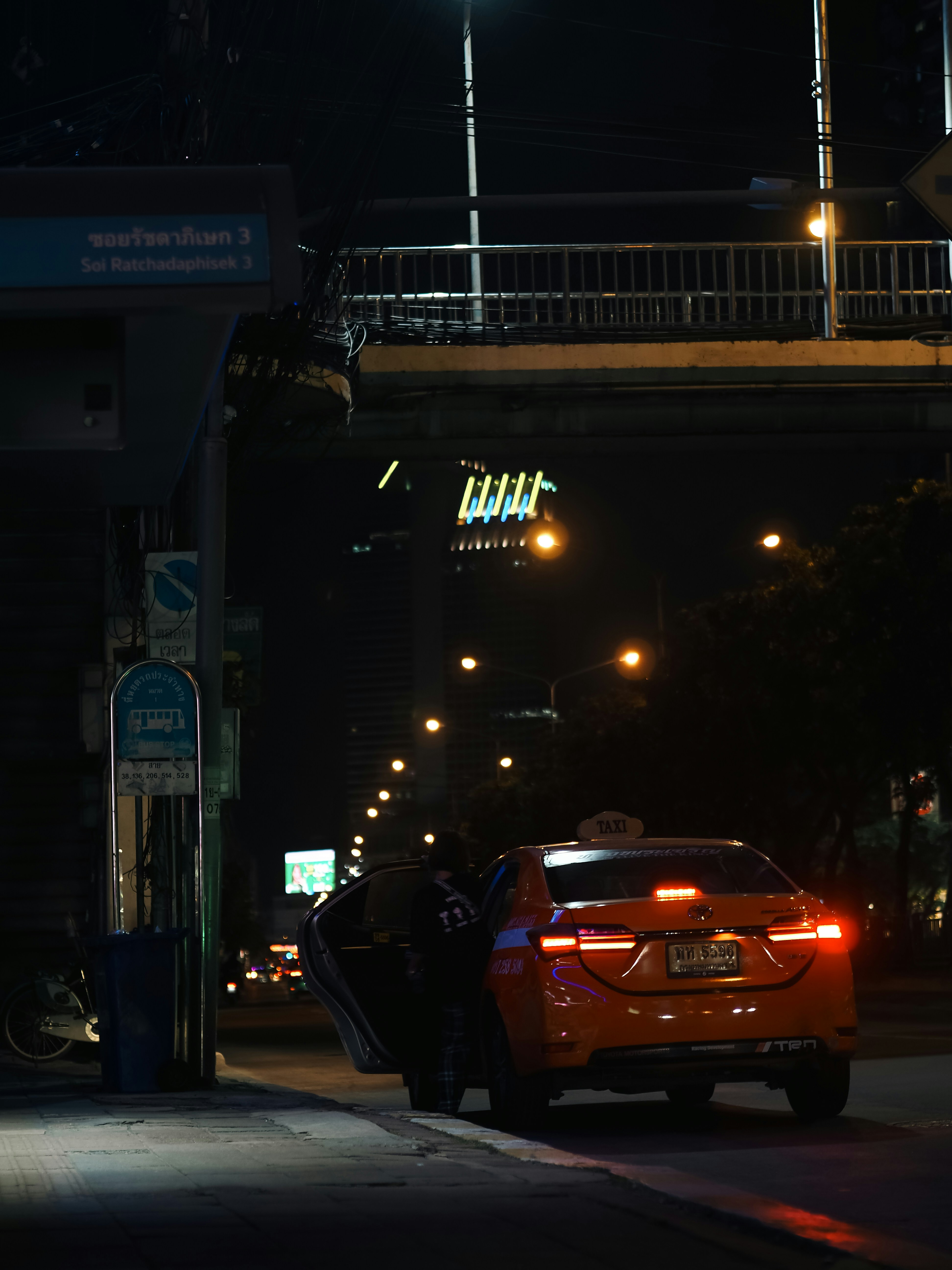 A brightly lit taxi waits on a bustling street corner, its door ajar as a passenger prepares to enter. Streetlights illuminate the scene, creating a vibrant city atmosphere.