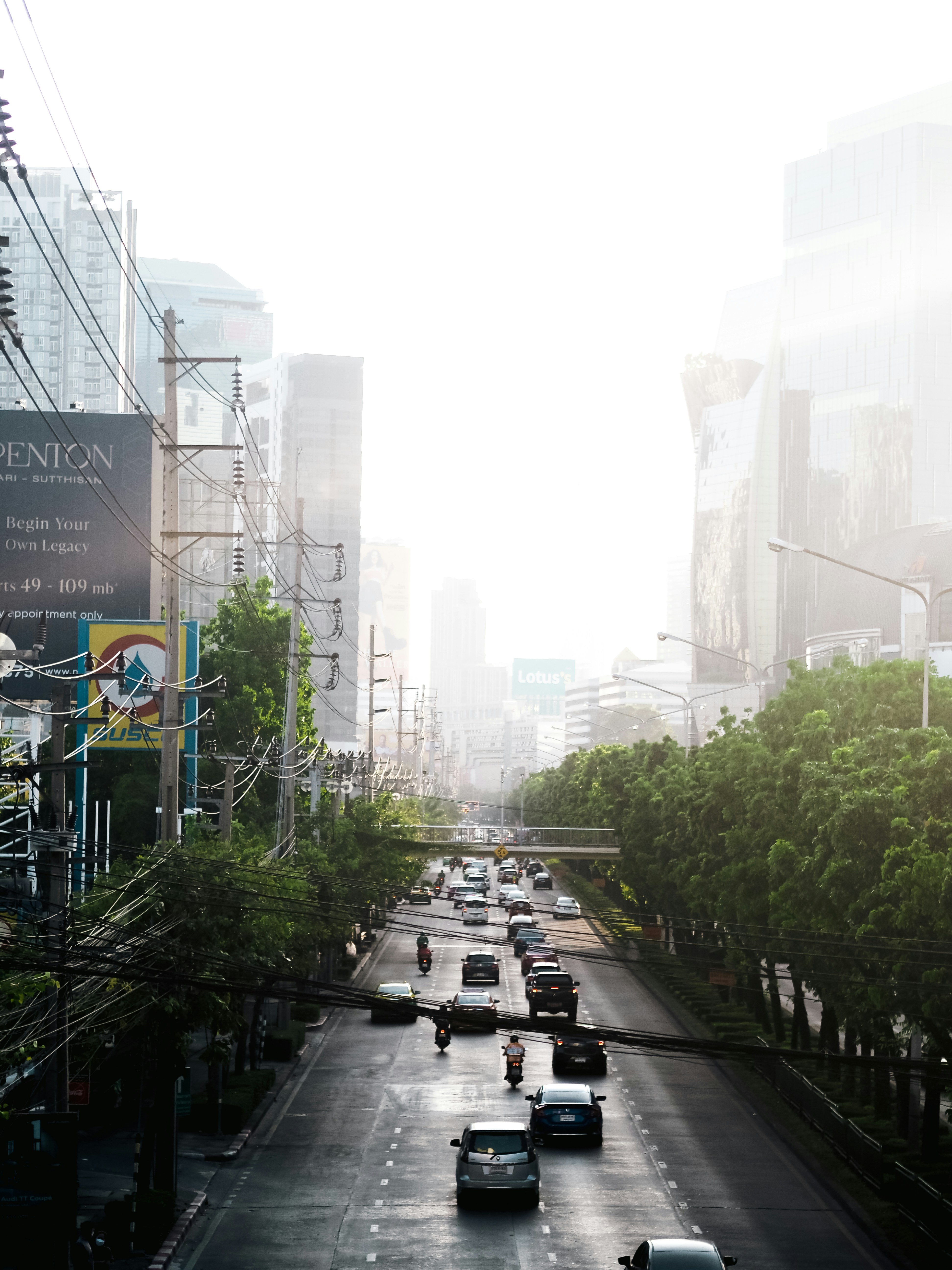 Rain-slick city avenue converges toward a bright skyline, with cars and motorcycles filling the lanes. Tall buildings, lush trees, and overhead power lines frame the scene.