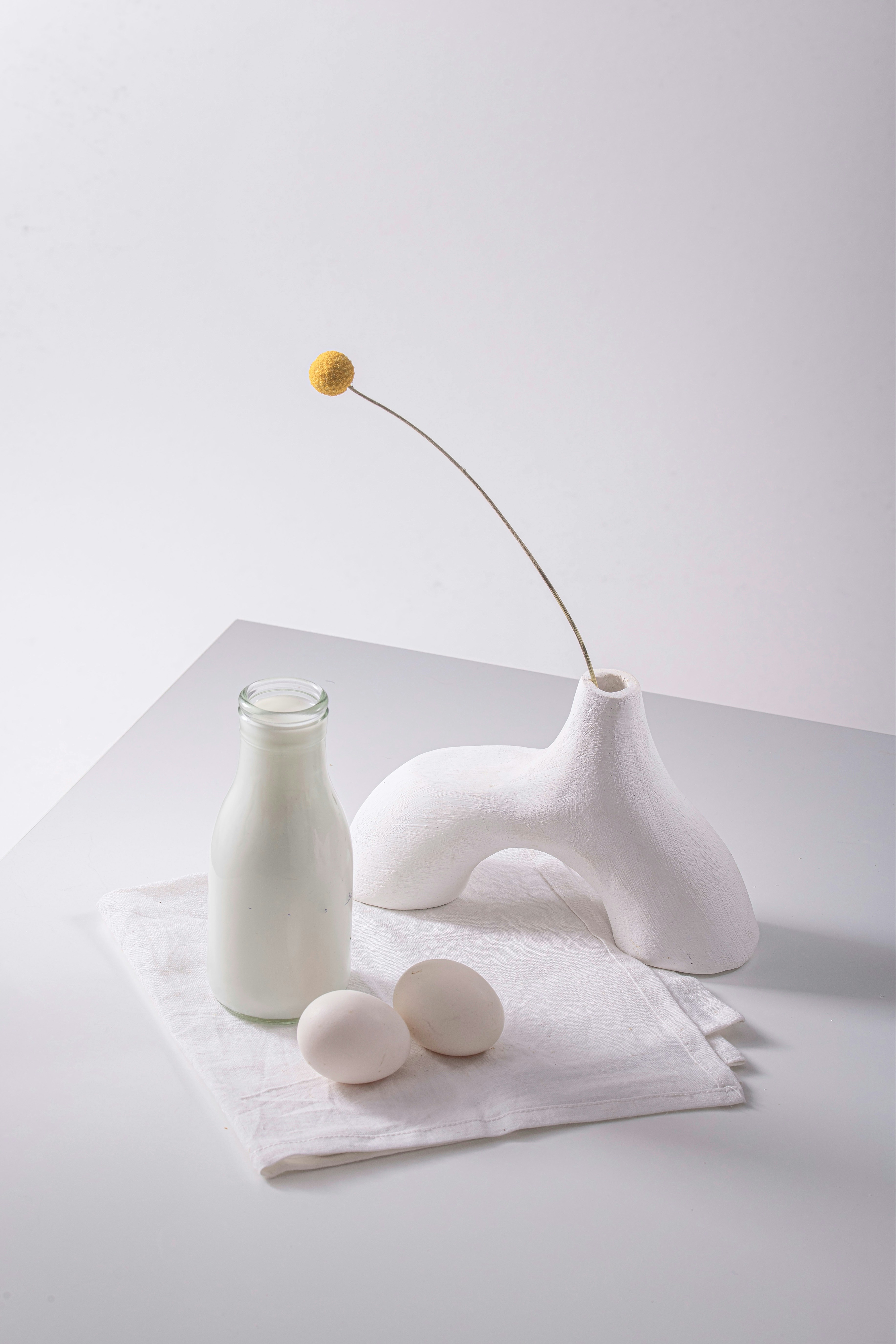 Arching Yellow Bloom Over a Minimal White Still Life: Milk Bottle, Eggs, and a Curved Ceramic Form