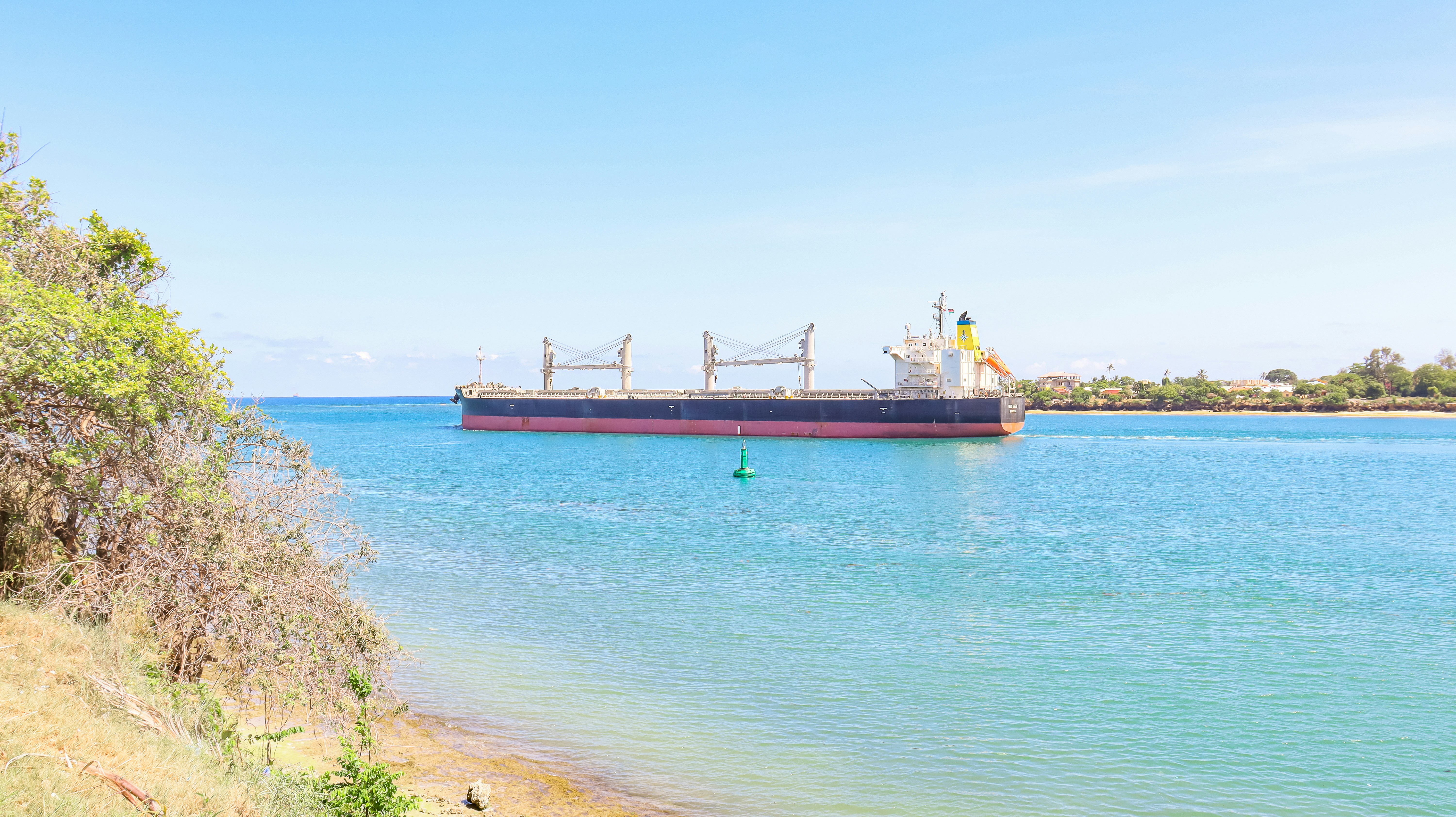 Large cargo ship navigating clear blue waters under a bright sky, with lush greenery on the shoreline.
