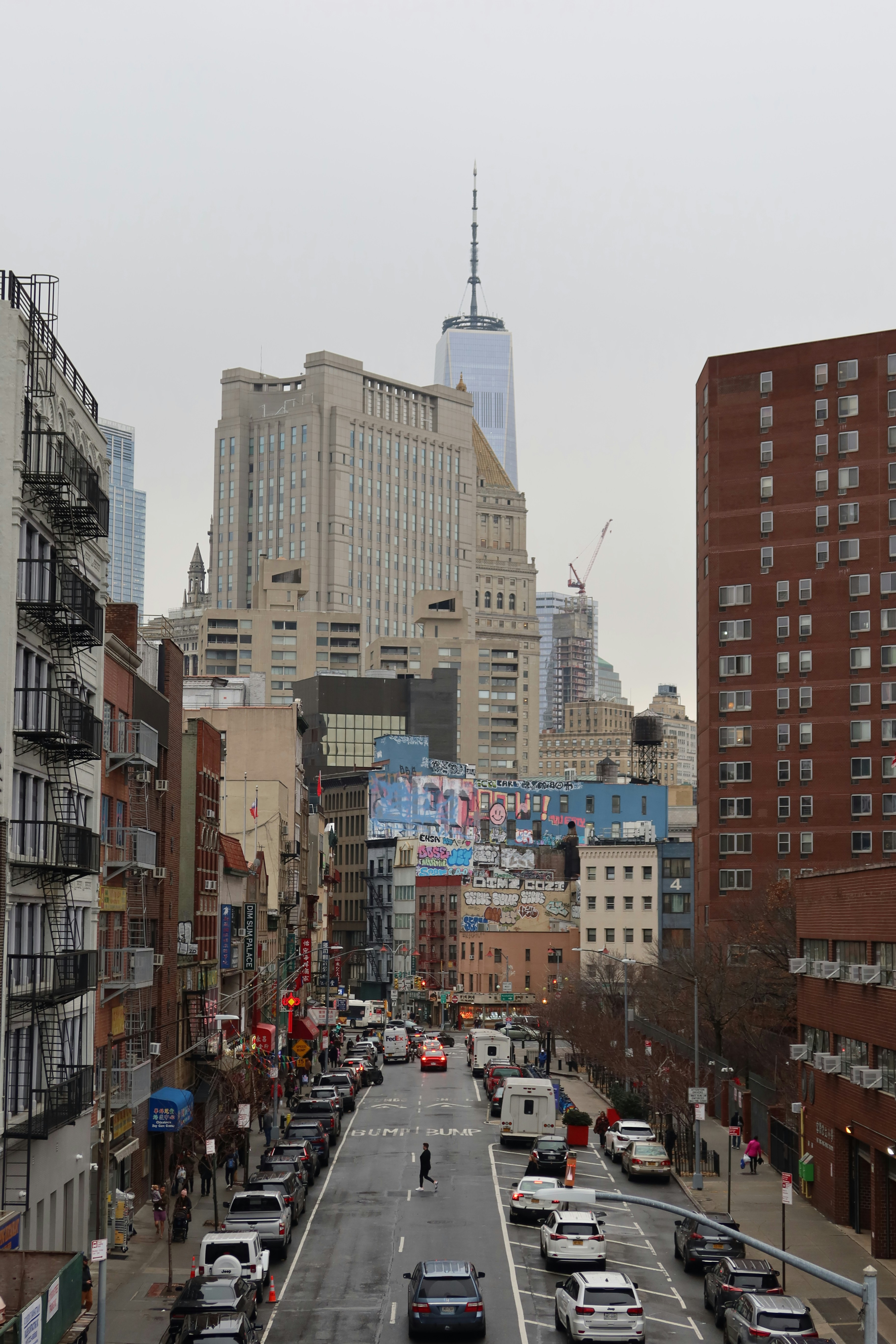 Bustling downtown street scene with mid-rise buildings framing a traffic-filled avenue, leading the eye to the One World Trade Center on the distant skyline.