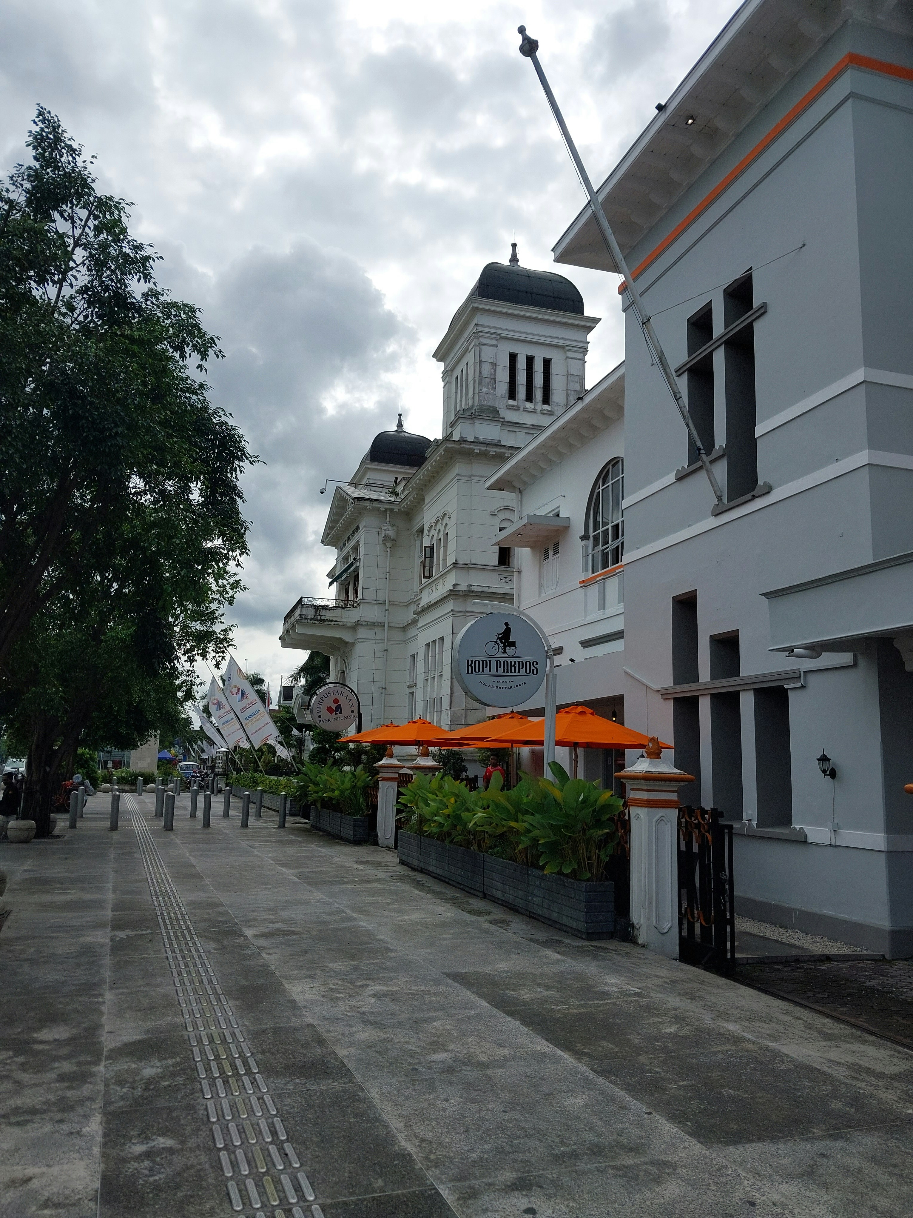 Historic building with ornate architecture and vibrant orange umbrellas lining the walkway. Lush greenery complements the urban setting.