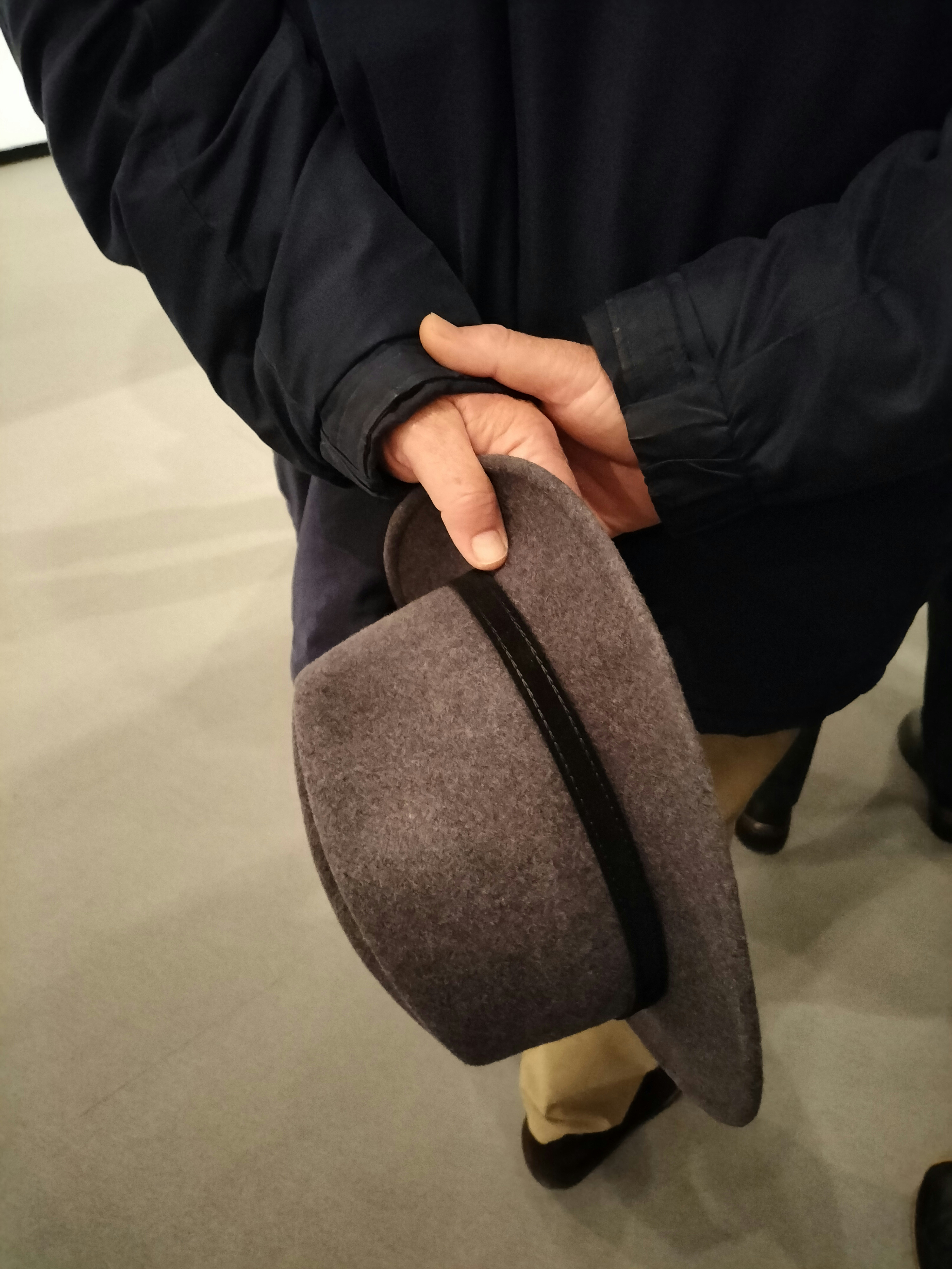 A photograph of two hands clasping a gray felt fedora with a black band, held against a dark jacket.