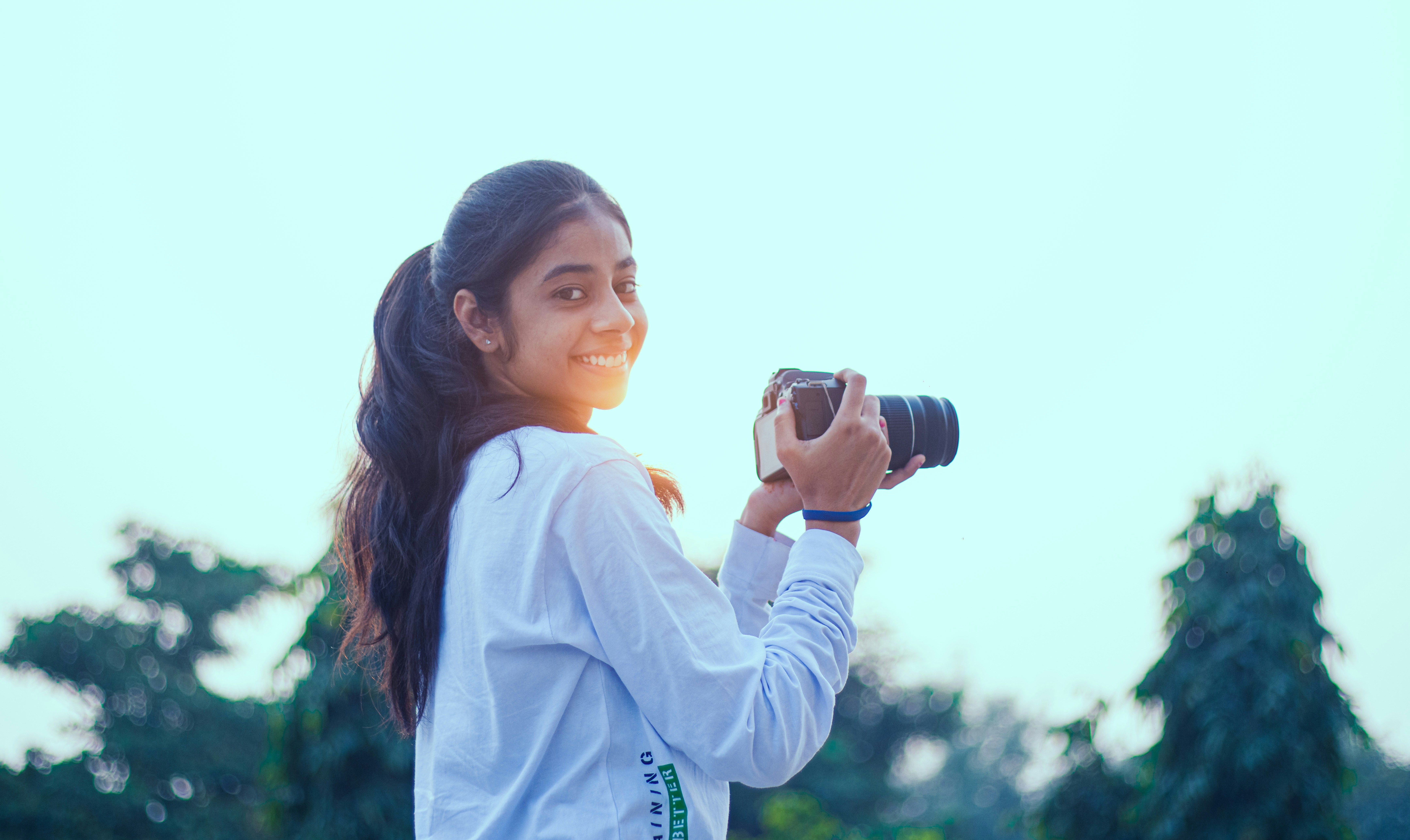 Young woman holding a camera, smiling in a lush outdoor setting during twilight.