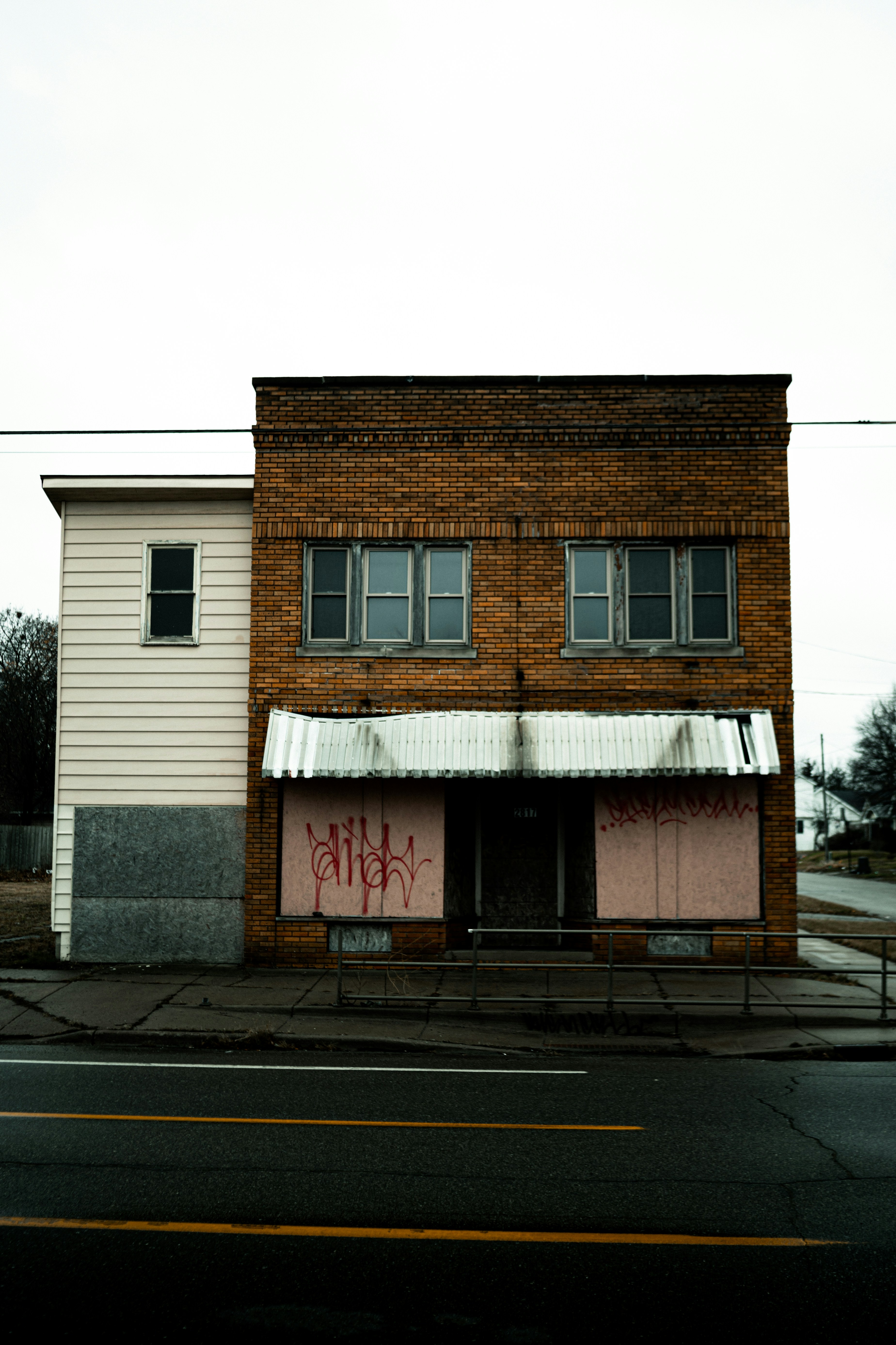 An aged building with a blend of brick and siding, featuring graffiti and a worn awning, set against a gray sky. The structure reflects urban decay and forgotten stories.