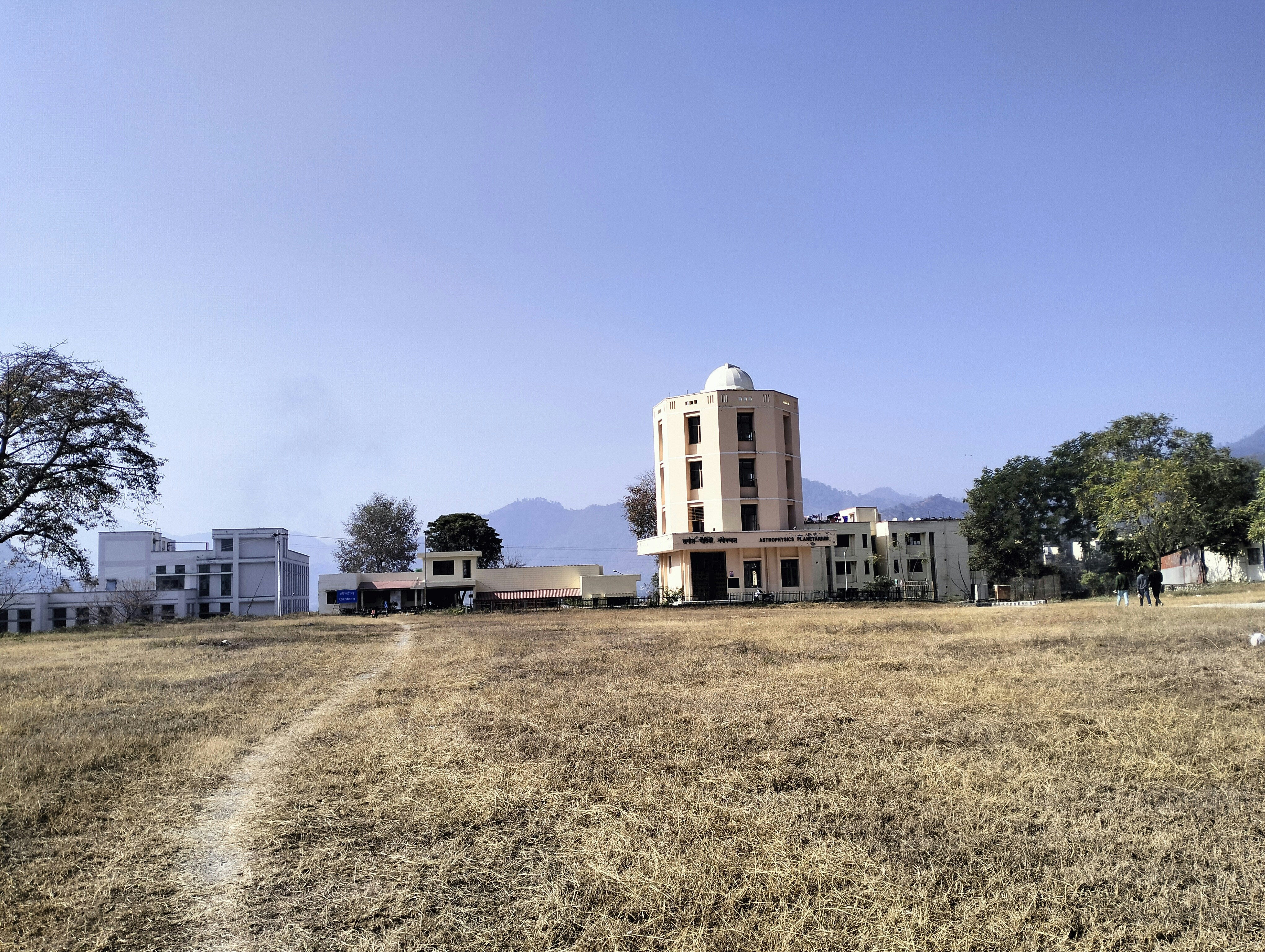 An empty, overgrown field with a prominent building in the center, surrounded by distant mountains and trees. The scene evokes a sense of nostalgia and stillness.