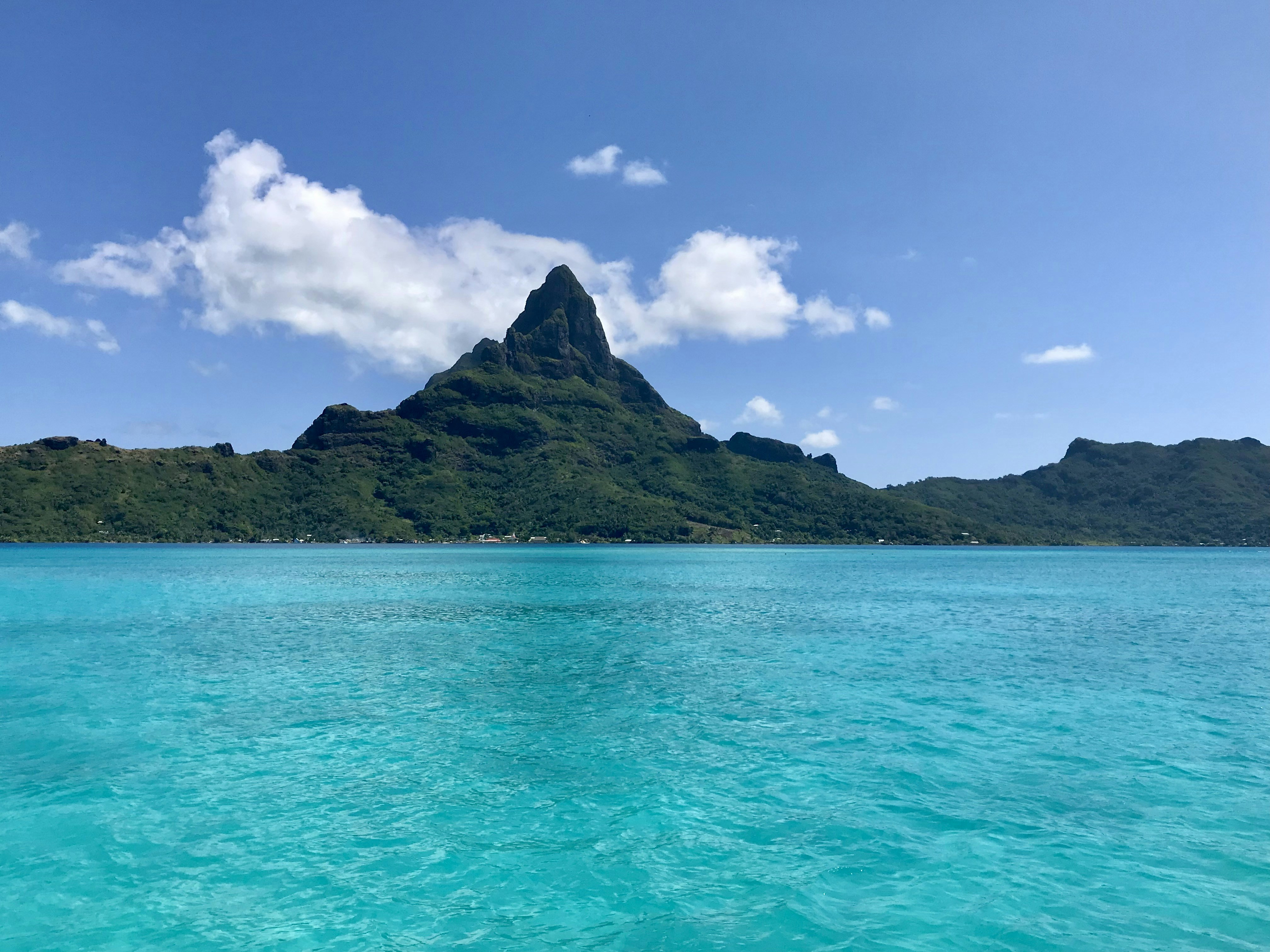 Lush green mountain rises dramatically from vibrant turquoise waters under a clear blue sky. Fluffy clouds drift above, enhancing the serene atmosphere.