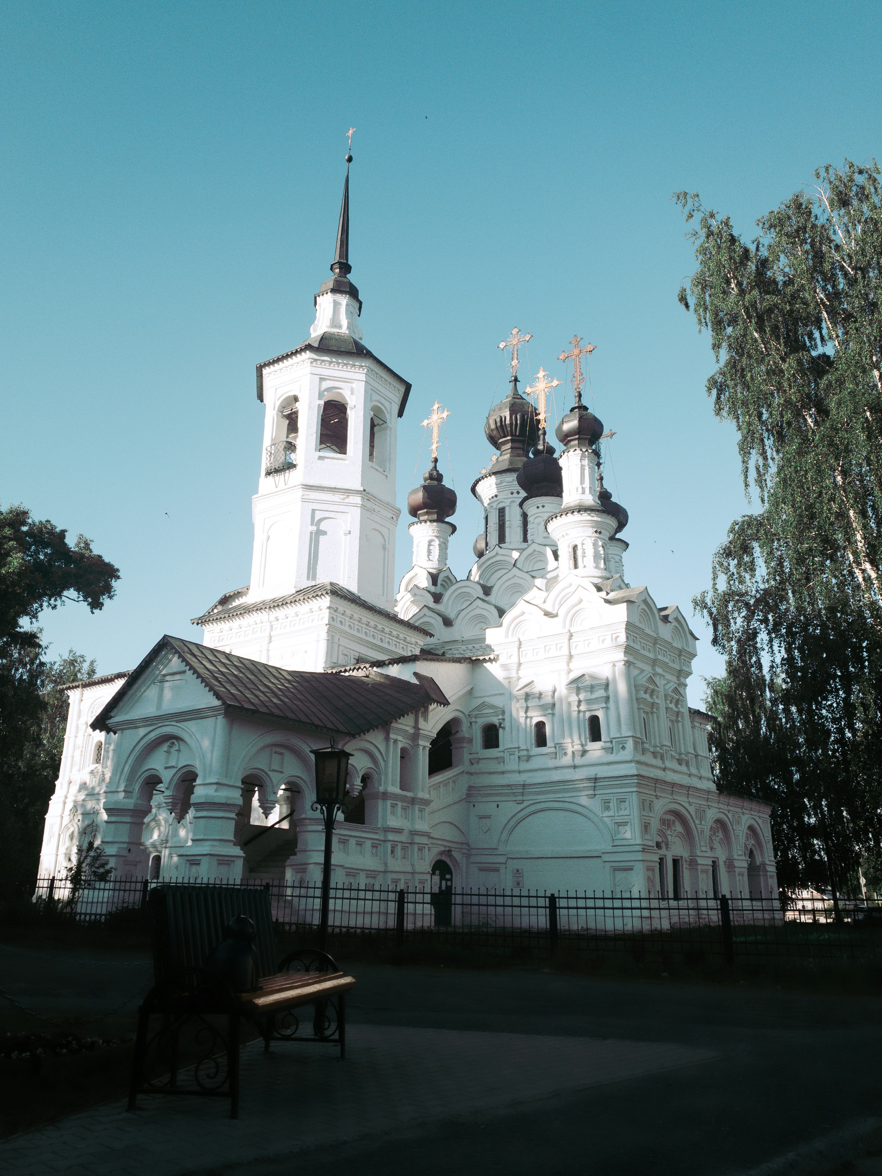 White church with ornate domes and crosses under a clear blue sky, flanked by lush green trees.