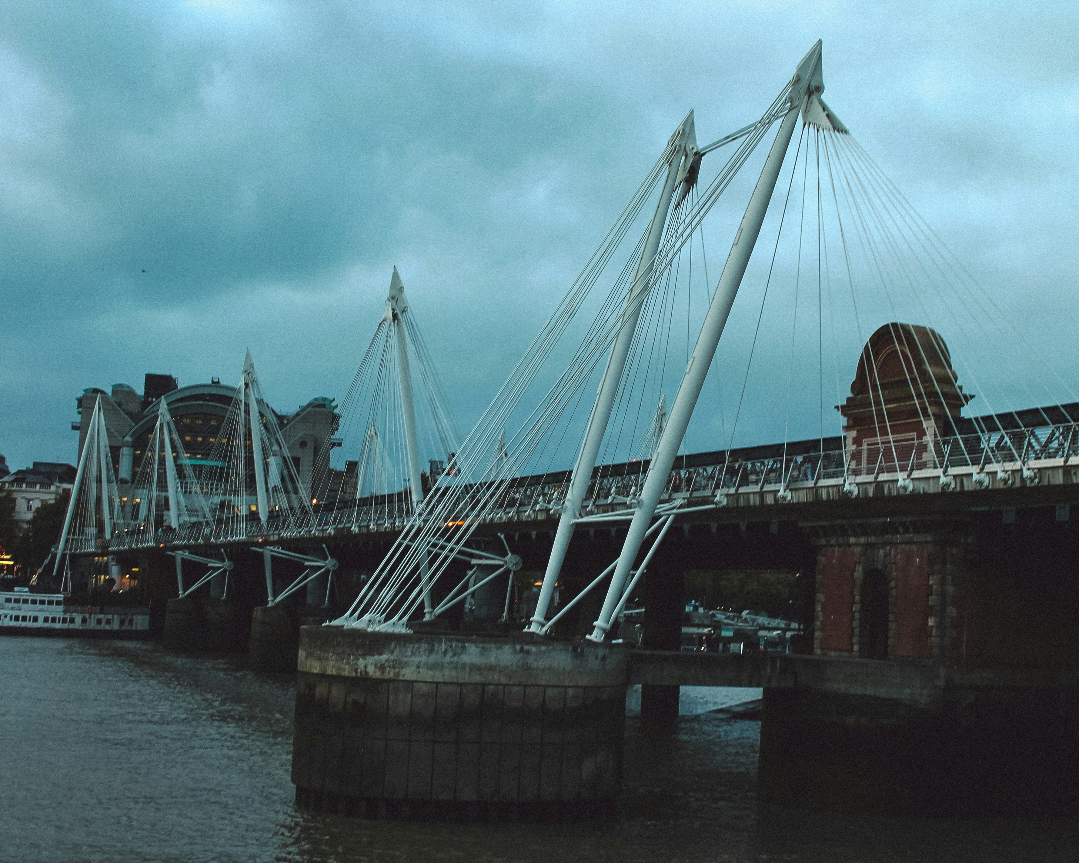 A cable-stayed bridge spans the Thames River, showcasing intricate architectural design against a moody sky. The scene captures the blend of urban infrastructure and natural beauty.