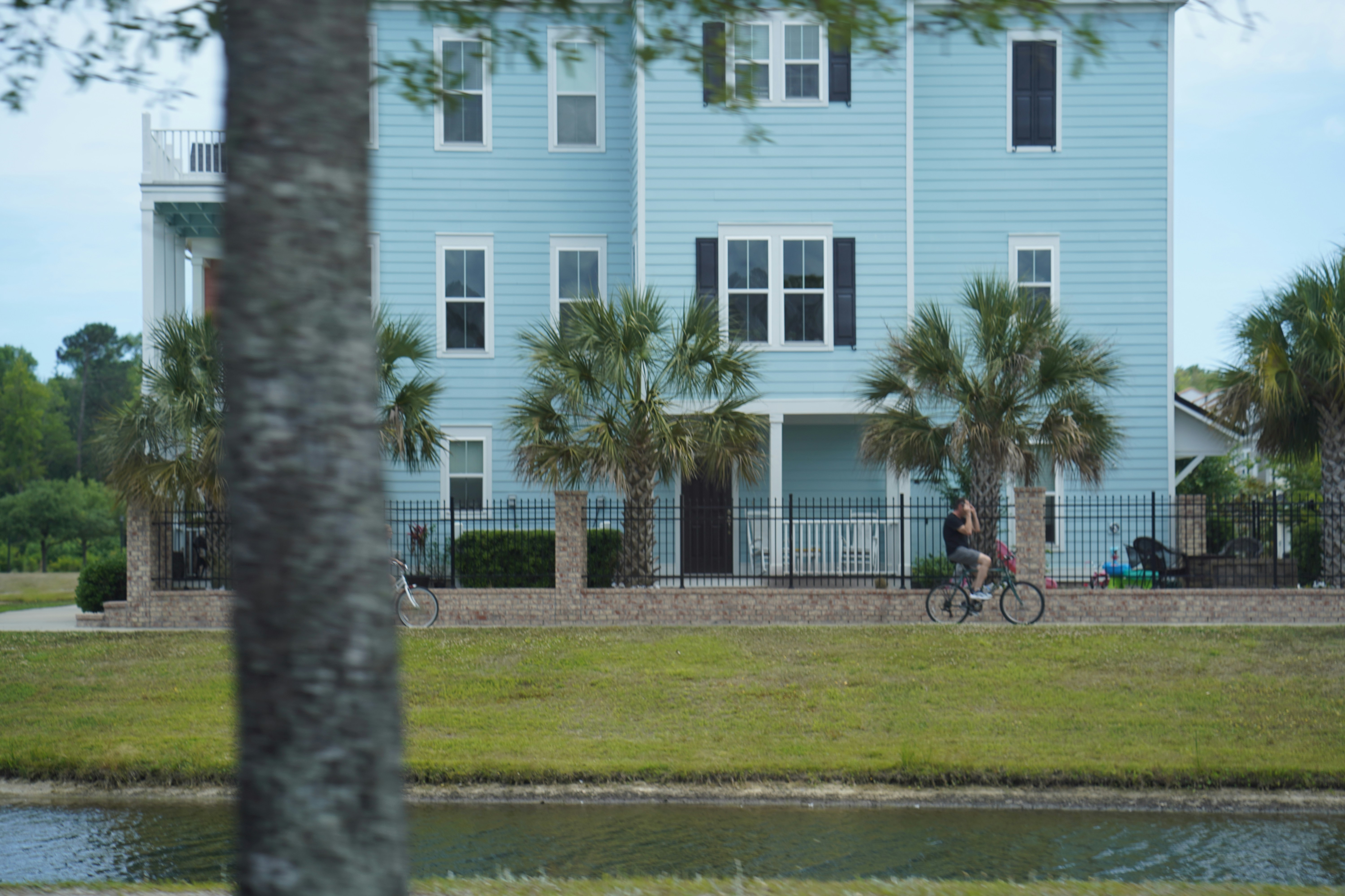 Cyclist riding past a large blue house with palm trees lining the entrance.