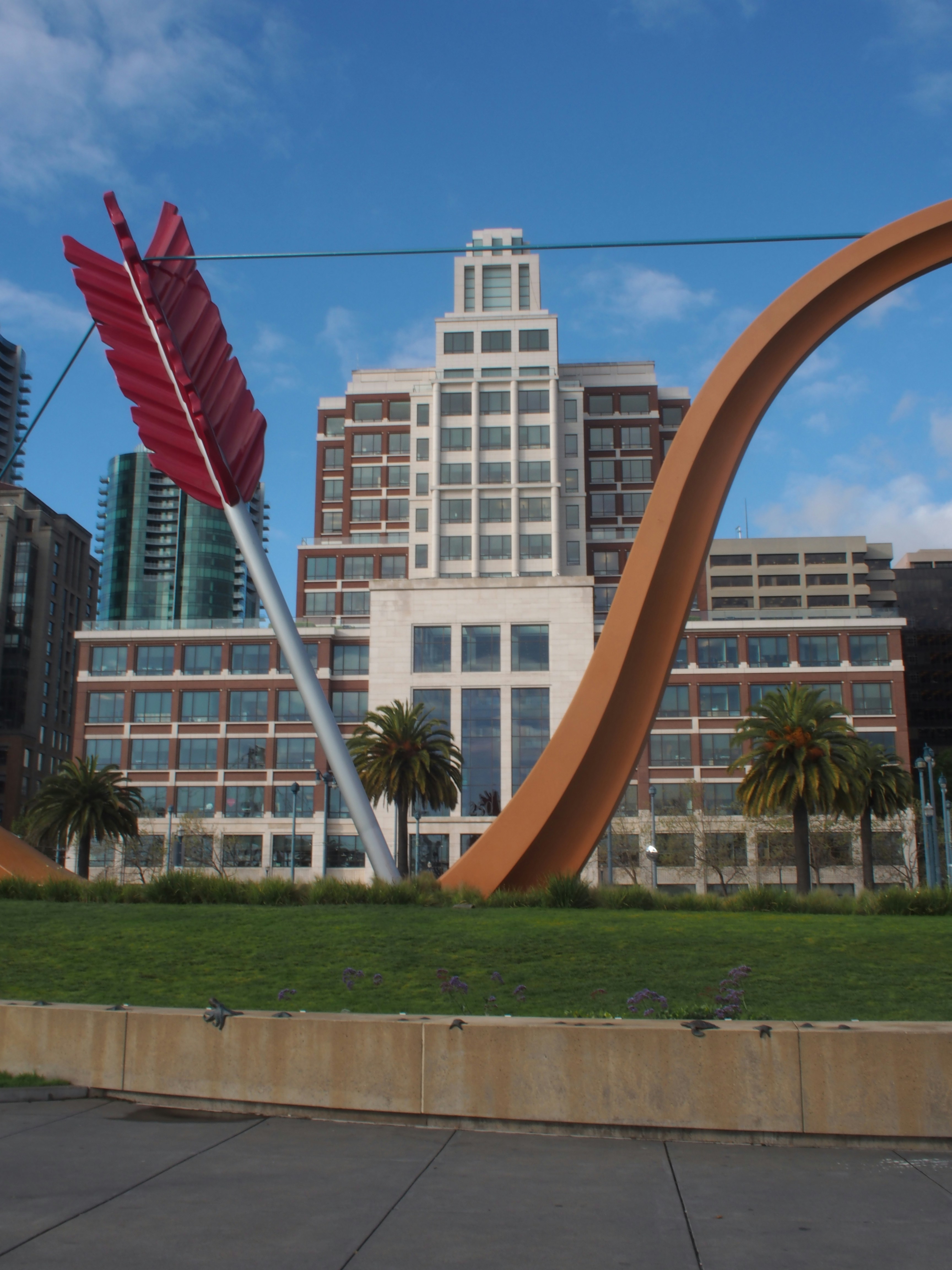 Outdoor sculpture installation in an urban plaza featuring a large red arrow and a sweeping orange arch set against a neo‑Art Deco building; palm trees and a manicured lawn anchor the foreground.