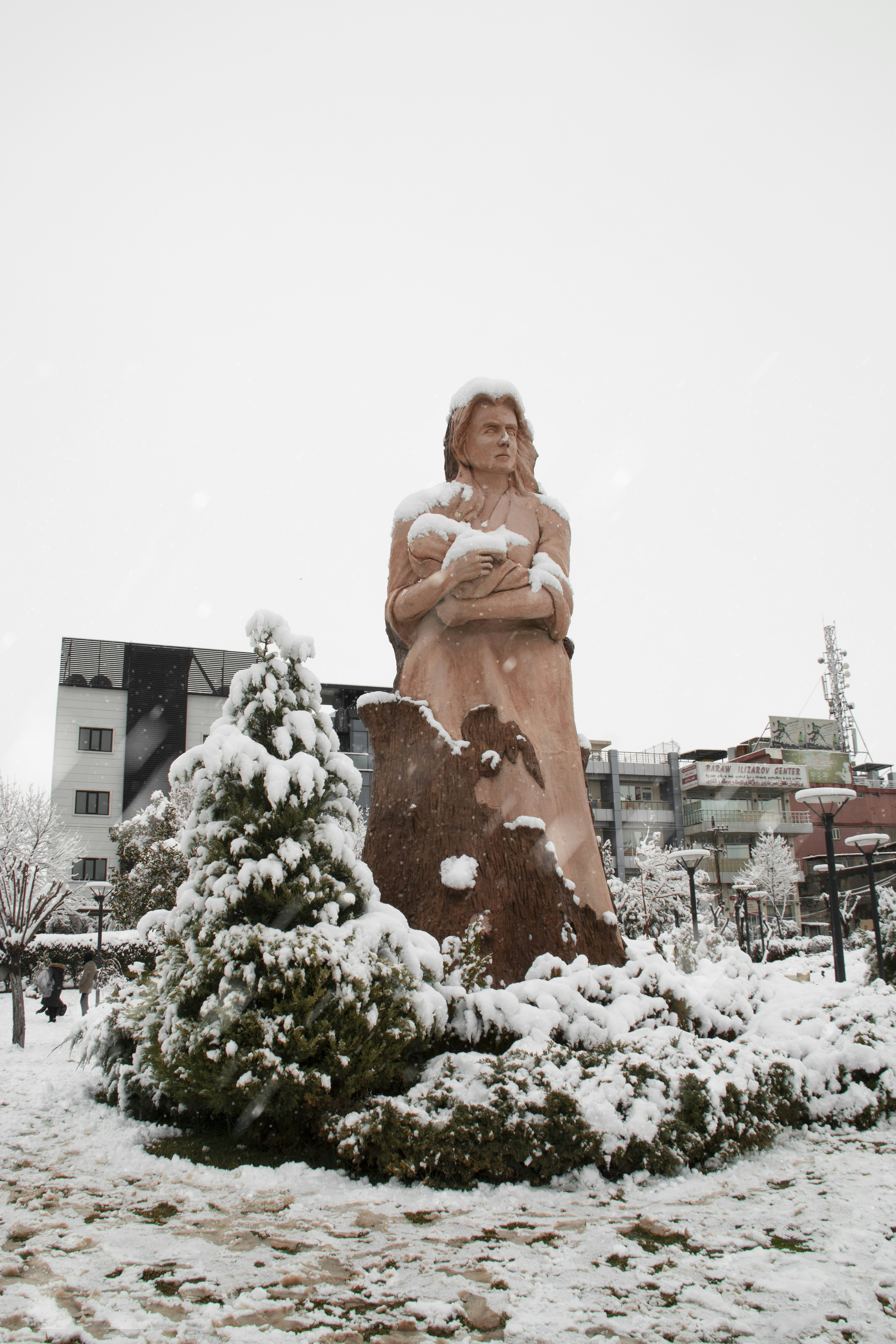 A towering statue of a woman cradling a child, blanketed in fresh snow, surrounded by snow-dusted pine trees in a serene park setting.