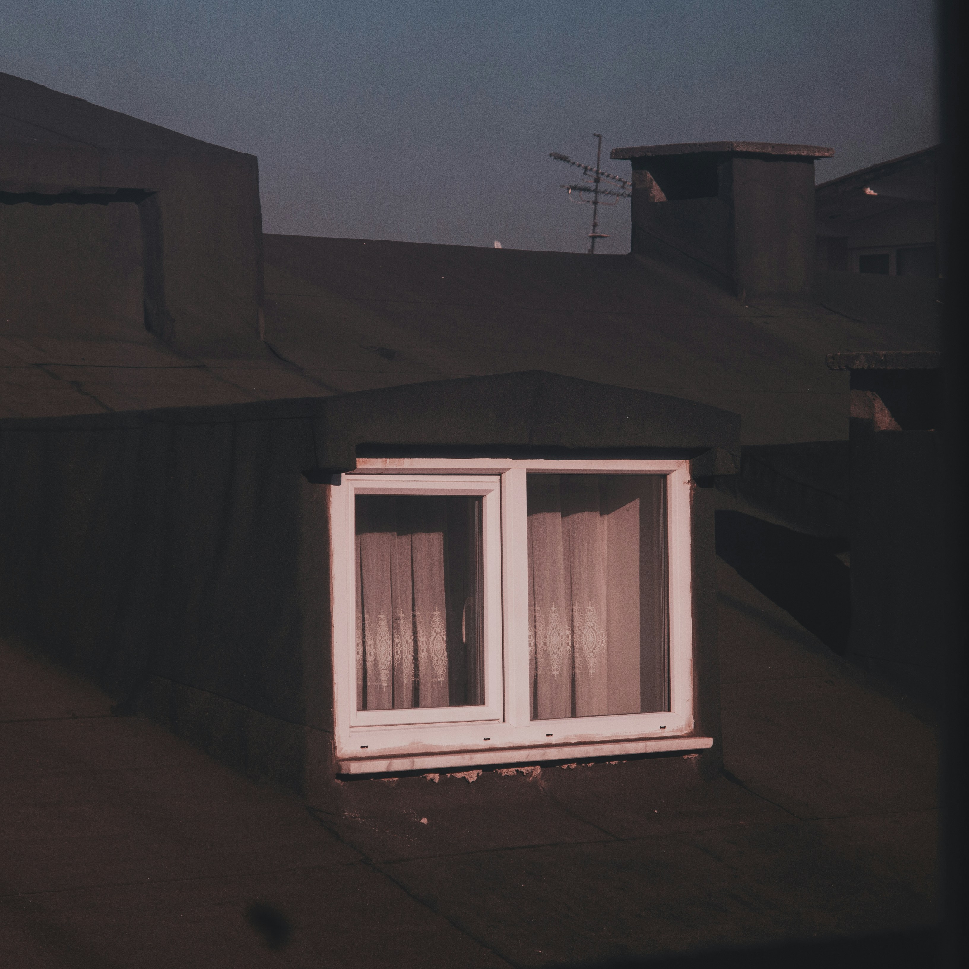 A sunlit window framed by dark rooftops, capturing the contrast of urban architecture. The scene evokes a sense of solitude and introspection.