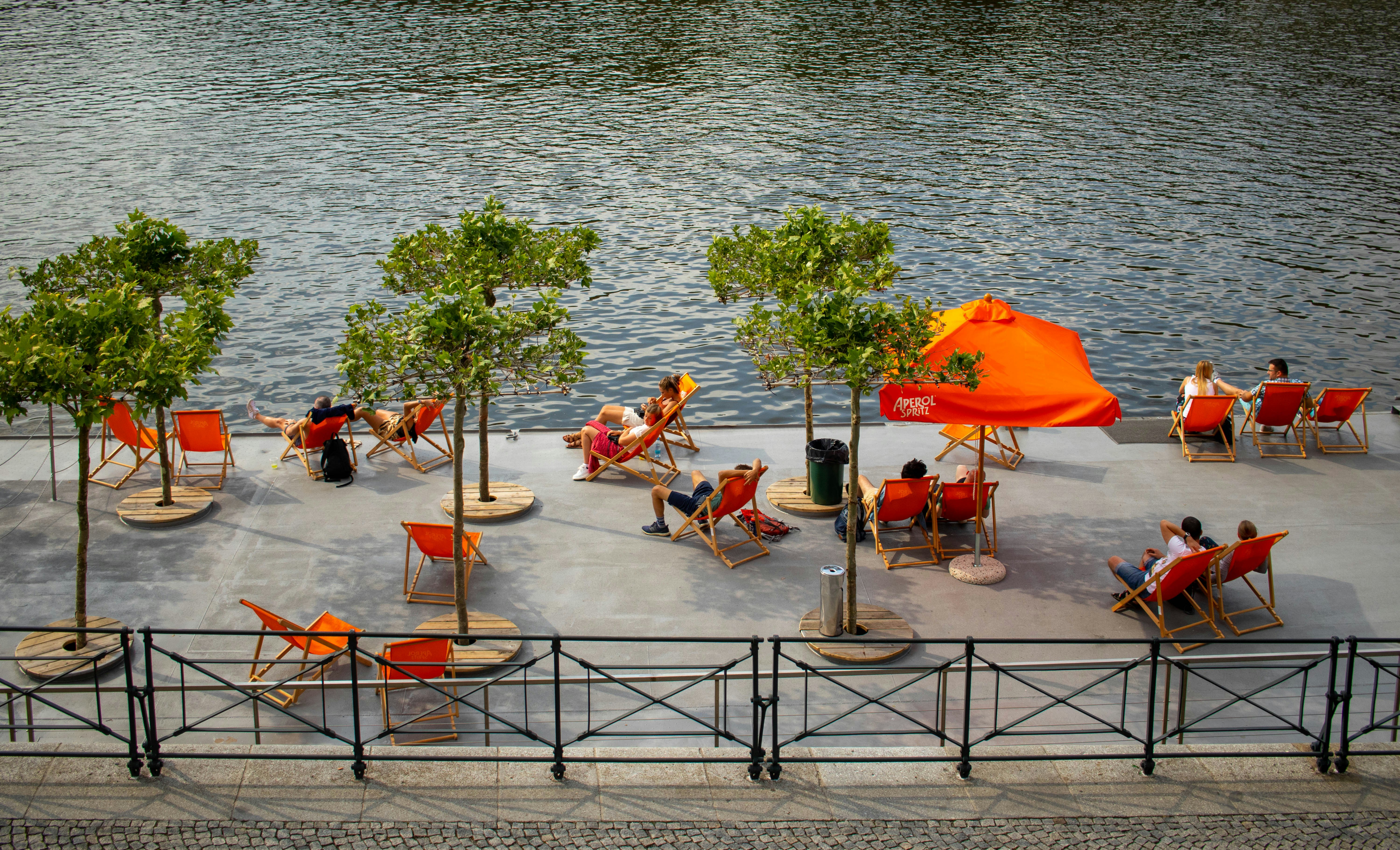 Sunlit Riverside Retreat: Orange Umbrellas and Loungers along the Waterfront