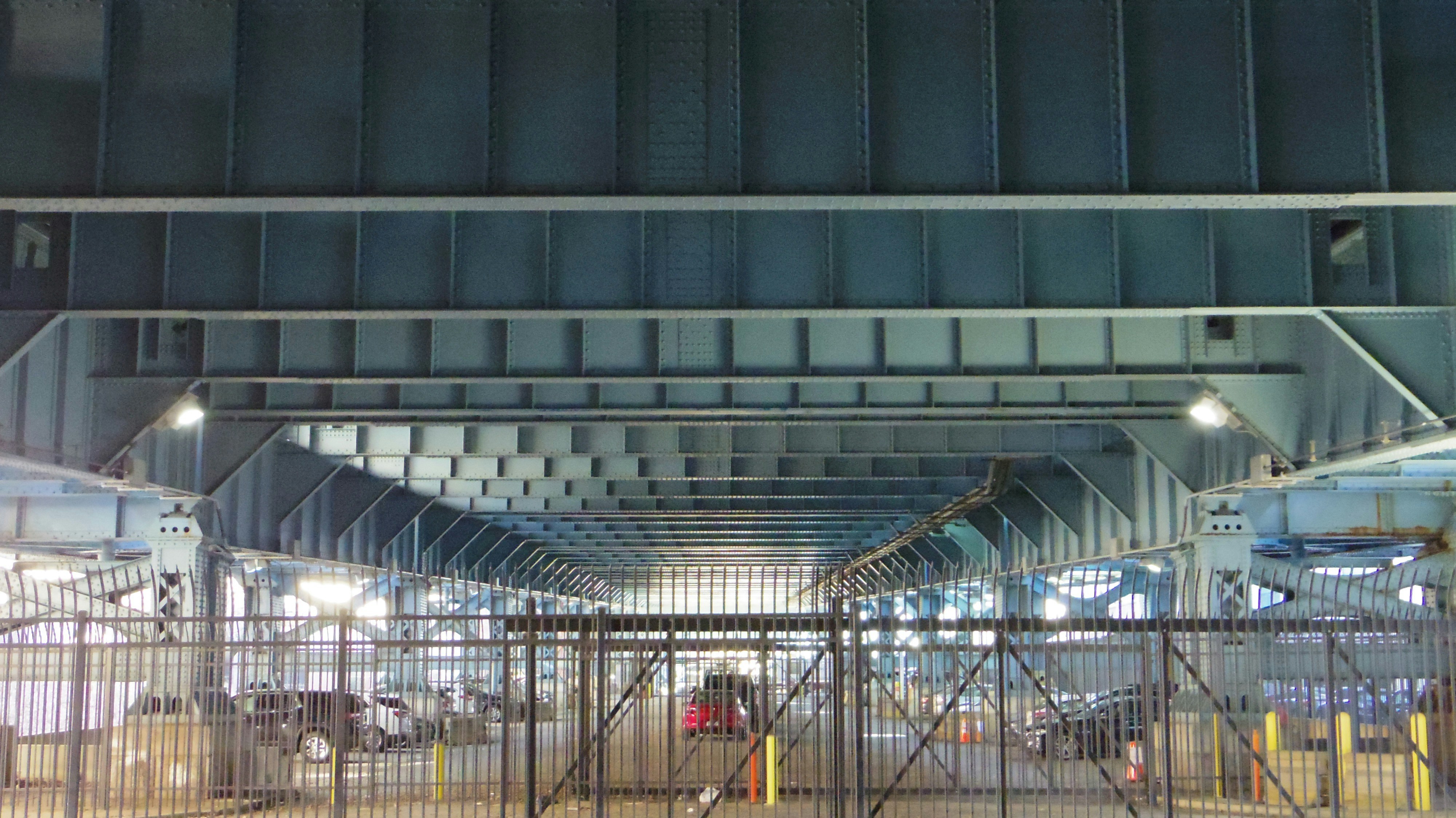 Industrial warehouse interior with repeating steel beams and bright overhead lighting. A red-clad figure stands at the far end of the central aisle.