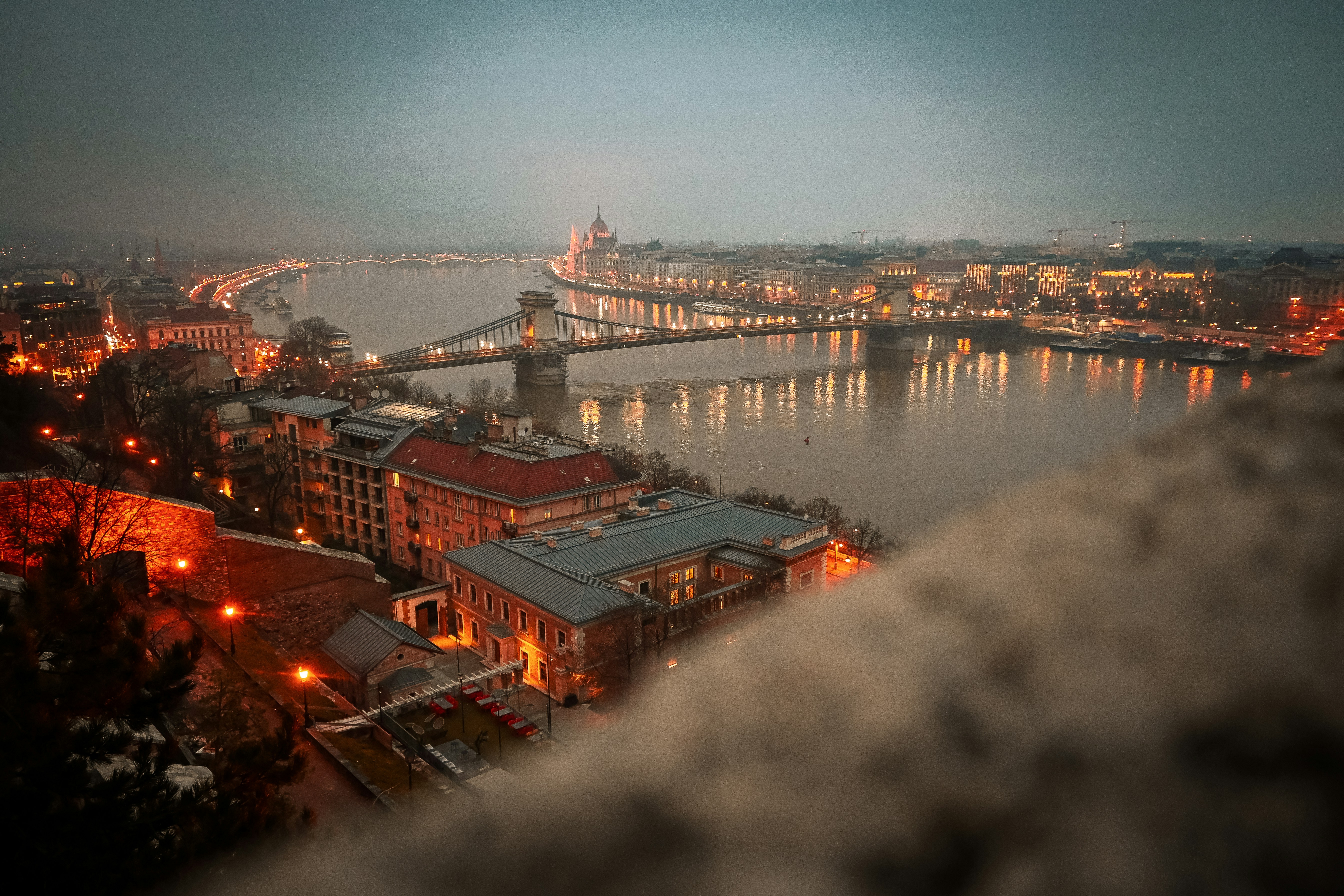 Snow-dusted foreground frames a lit riverside city with a glowing bridge spanning the Danube and reflections dancing on the water.