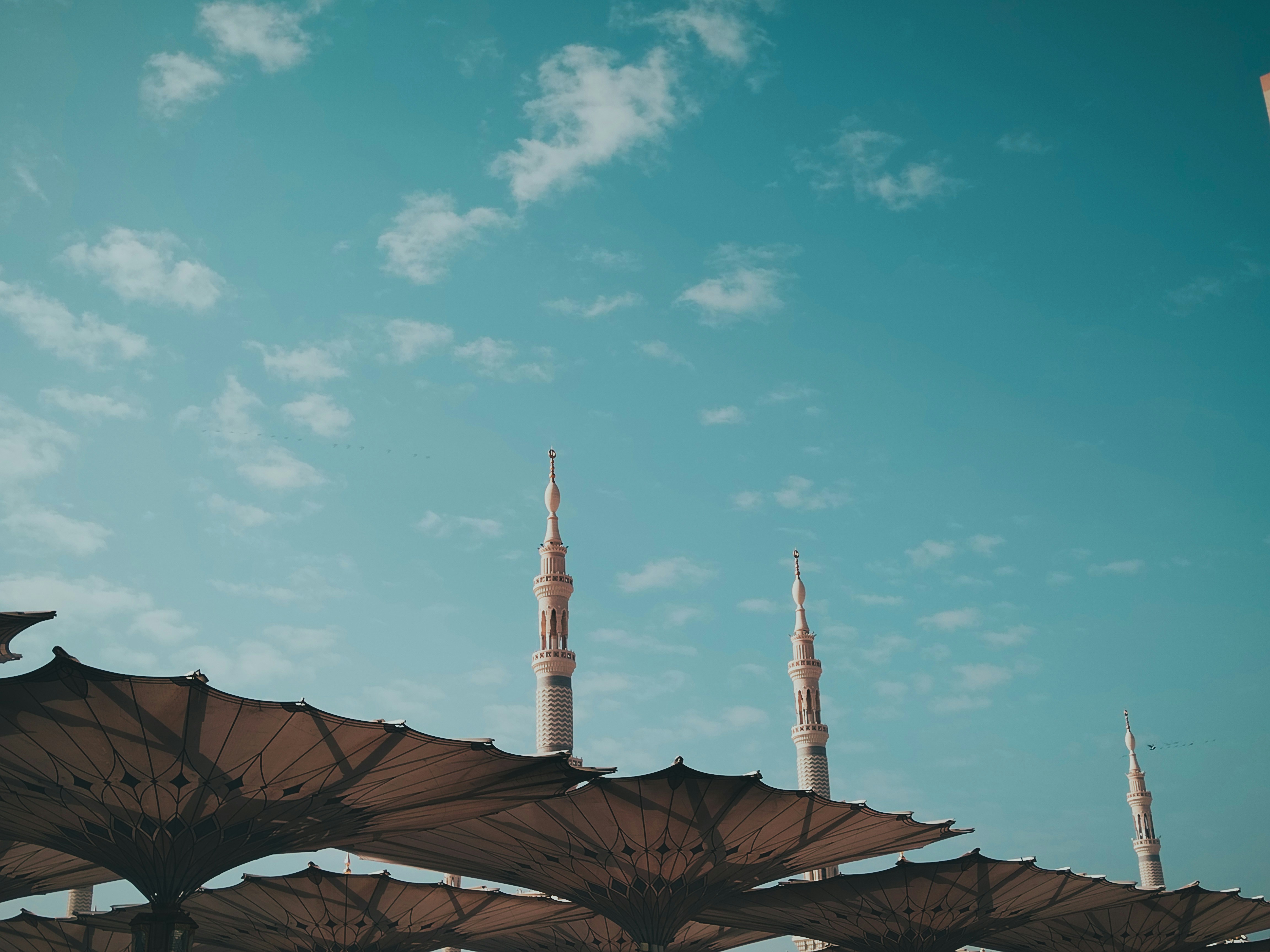 Blue sky with wispy clouds frames slender minarets rising above a distant cityscape, while large parasols in the foreground create bold geometric silhouettes.