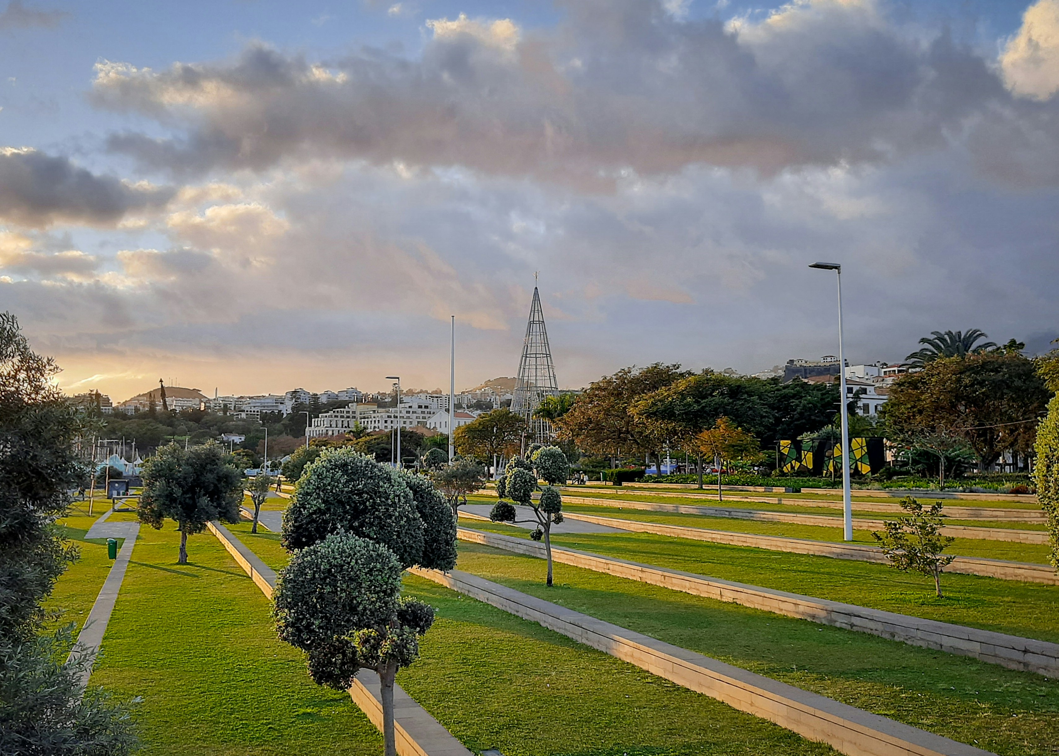 Wide-angle photograph of a manicured park at dusk, featuring winding paths, sculpted shrubs, and a distant lattice tower on the horizon.