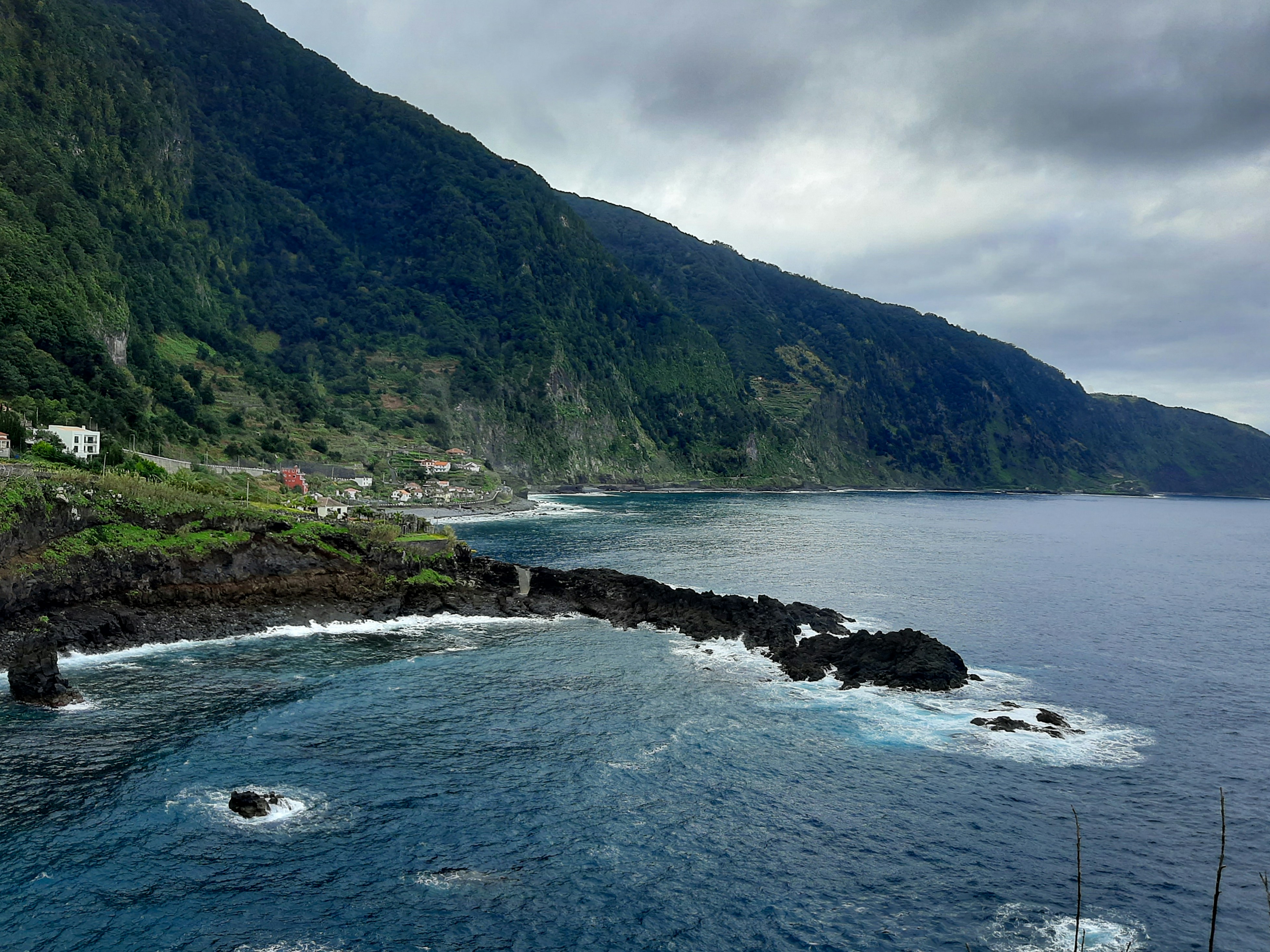 Lush green hills meet the rugged coastline, with waves crashing against dark rocks in a tranquil seascape. A small village nestles against the mountainside.