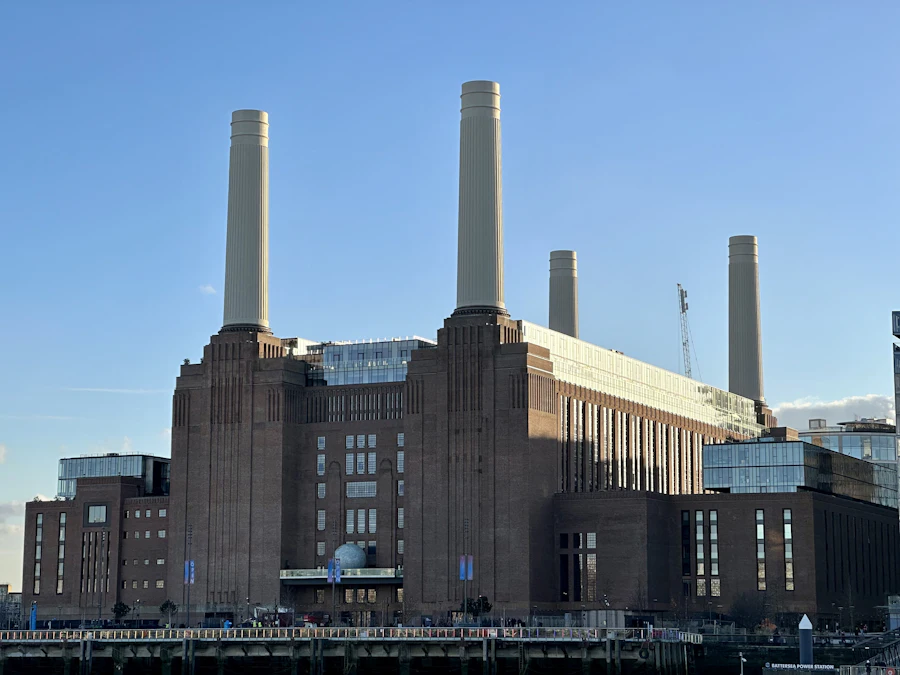 Exterior of Battersea Power Station with four tall chimneys against a clear blue sky, viewed from a riverside walkway.