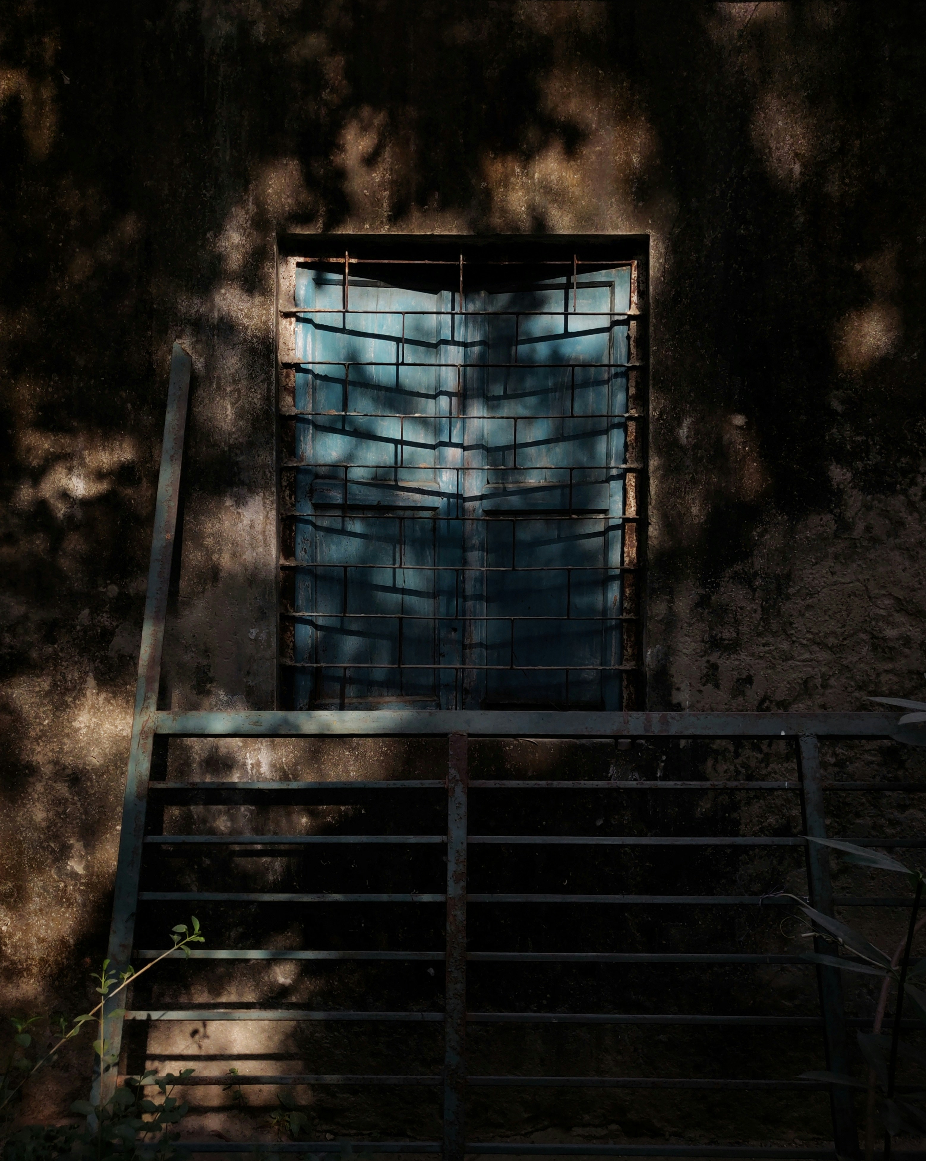 A weathered blue door behind a wrought iron grill, partially obscured by shadows and textured walls, evokes a sense of mystery. The interplay of light and dark adds depth to the scene.