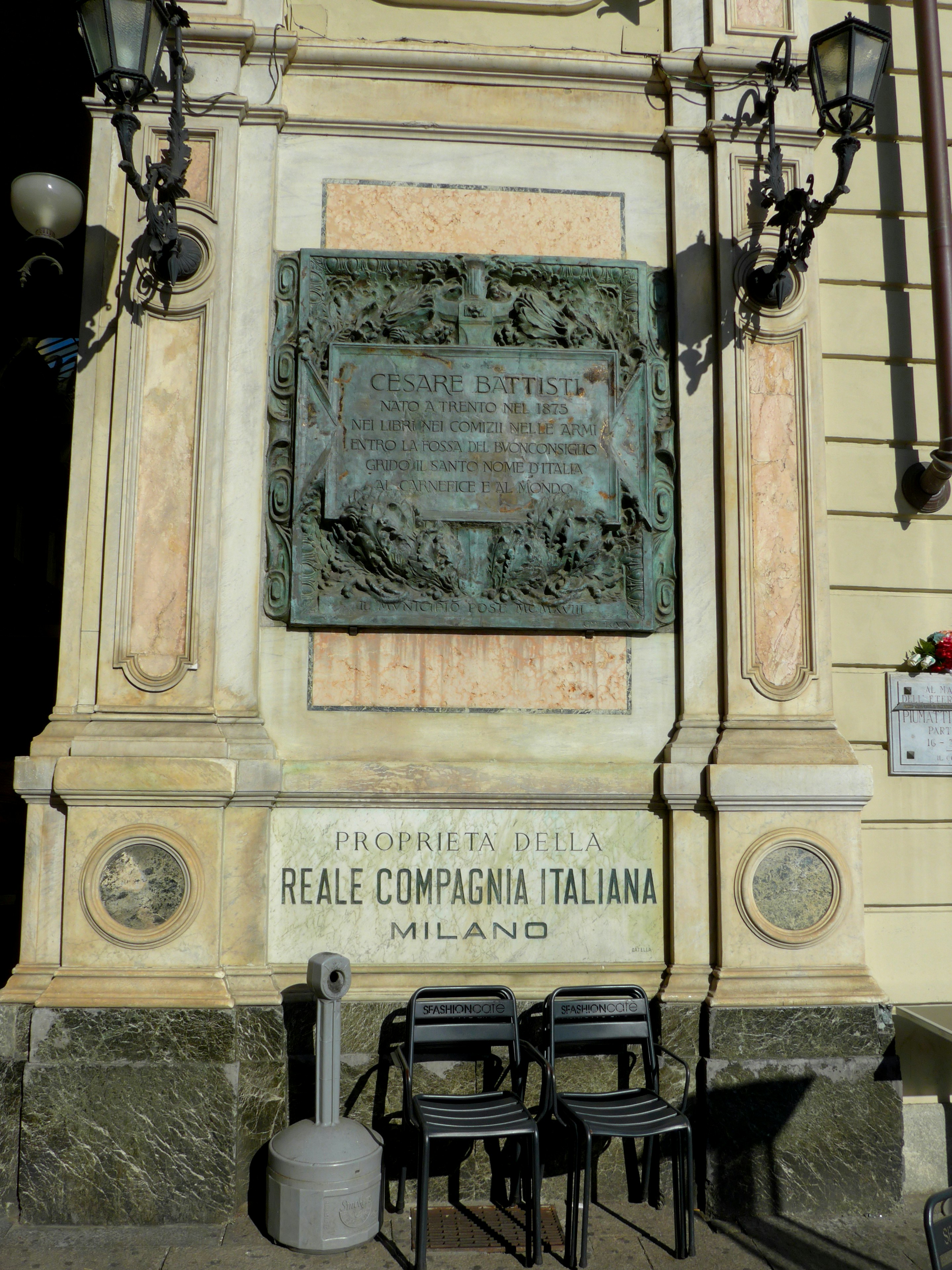 Photograph of a historic bronze plaque mounted on a Milan building façade, flanked by ornate lanterns and a stack of chairs in front.