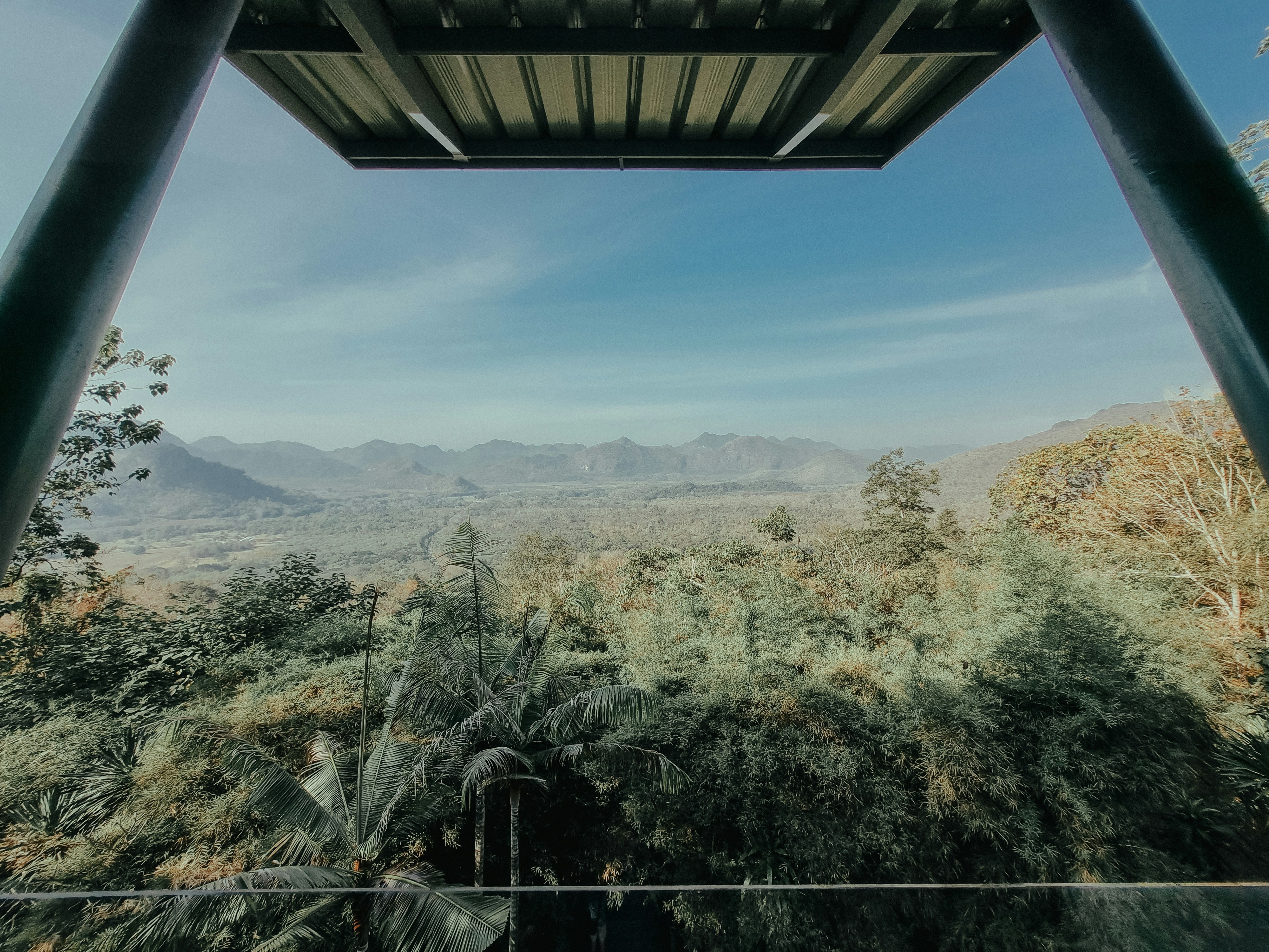 View from a modern structure overlooking dense green foliage and distant mountains under a clear blue sky.