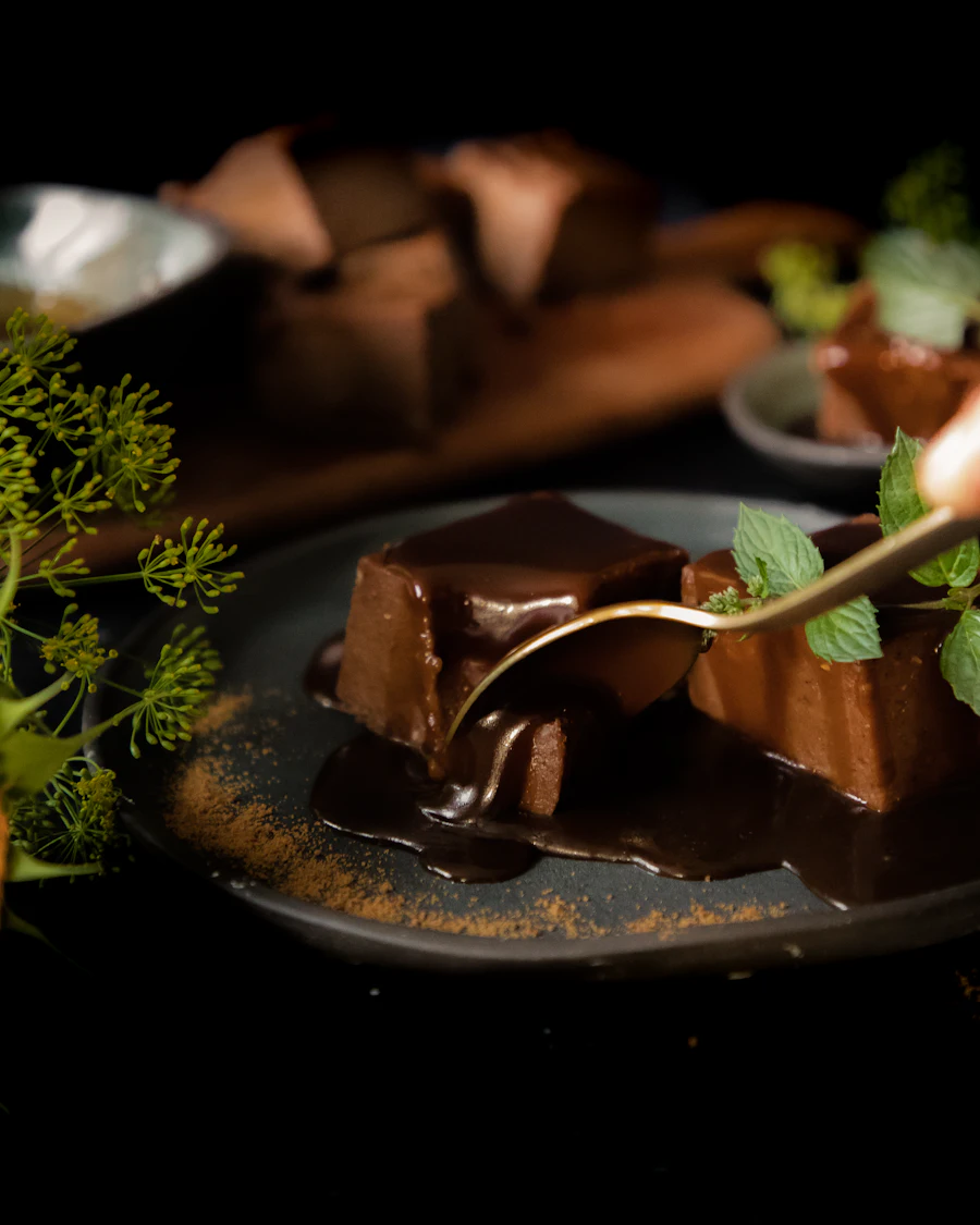 Plated chocolate dessert squares with glossy chocolate sauce, mint leaves, and a spoon lifting a bite. Dark, moody styling with herbs framing the plate.