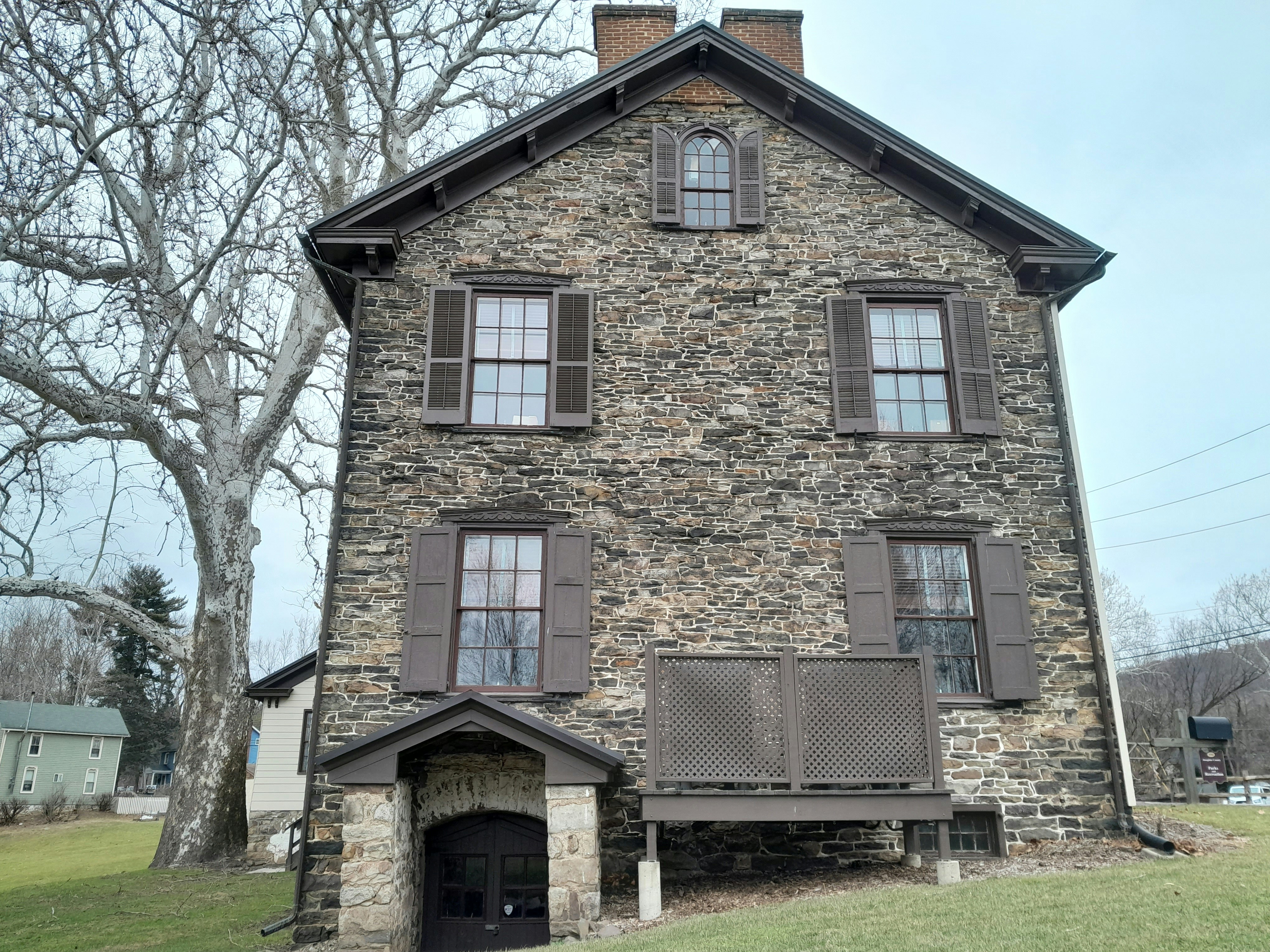 Historic stone house with wooden shutters and a distinctive porch, surrounded by trees and a grassy yard.