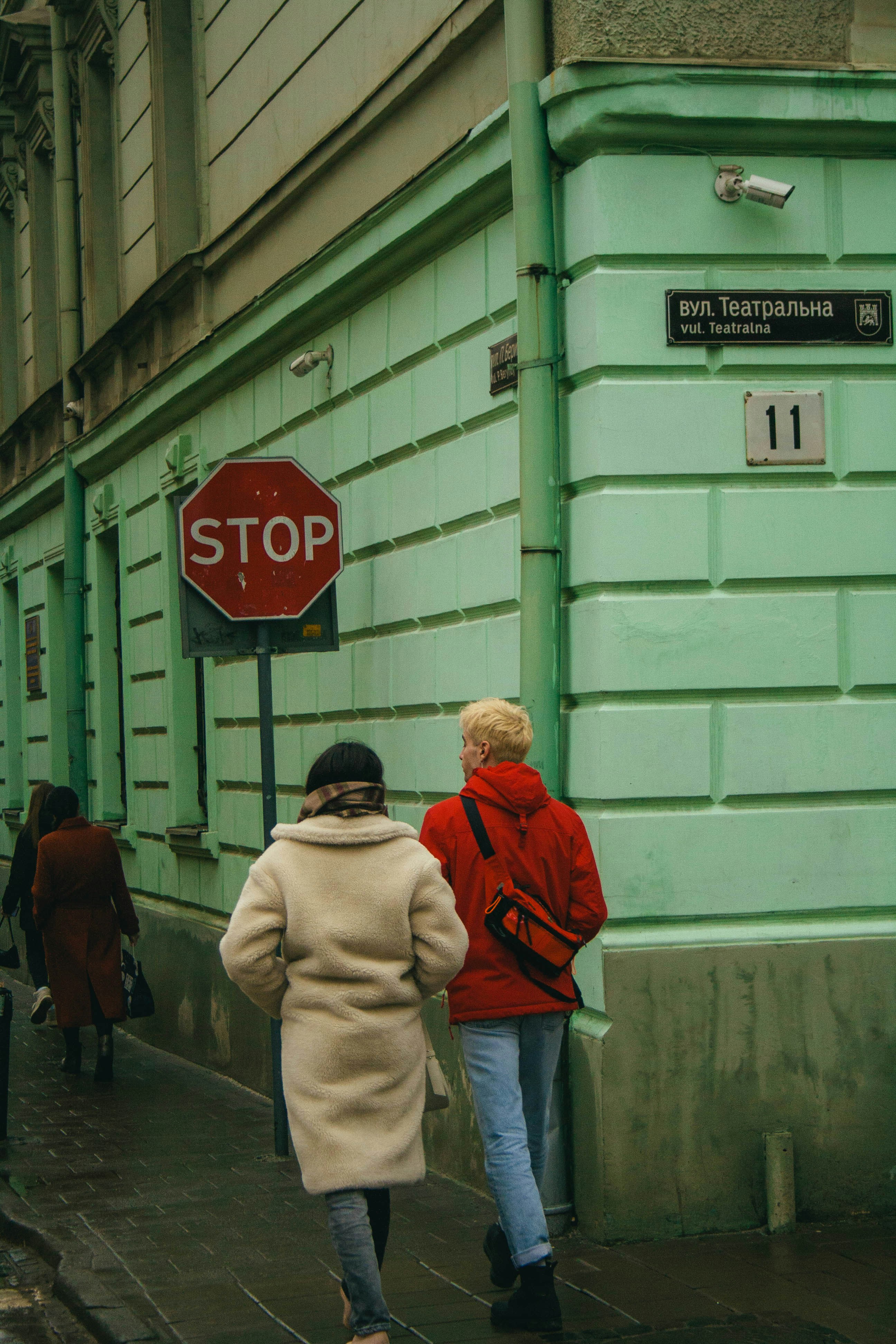 Two figures walking past a stop sign on a city street, framed by a pastel green building. The scene captures a blend of urban life and architectural detail.