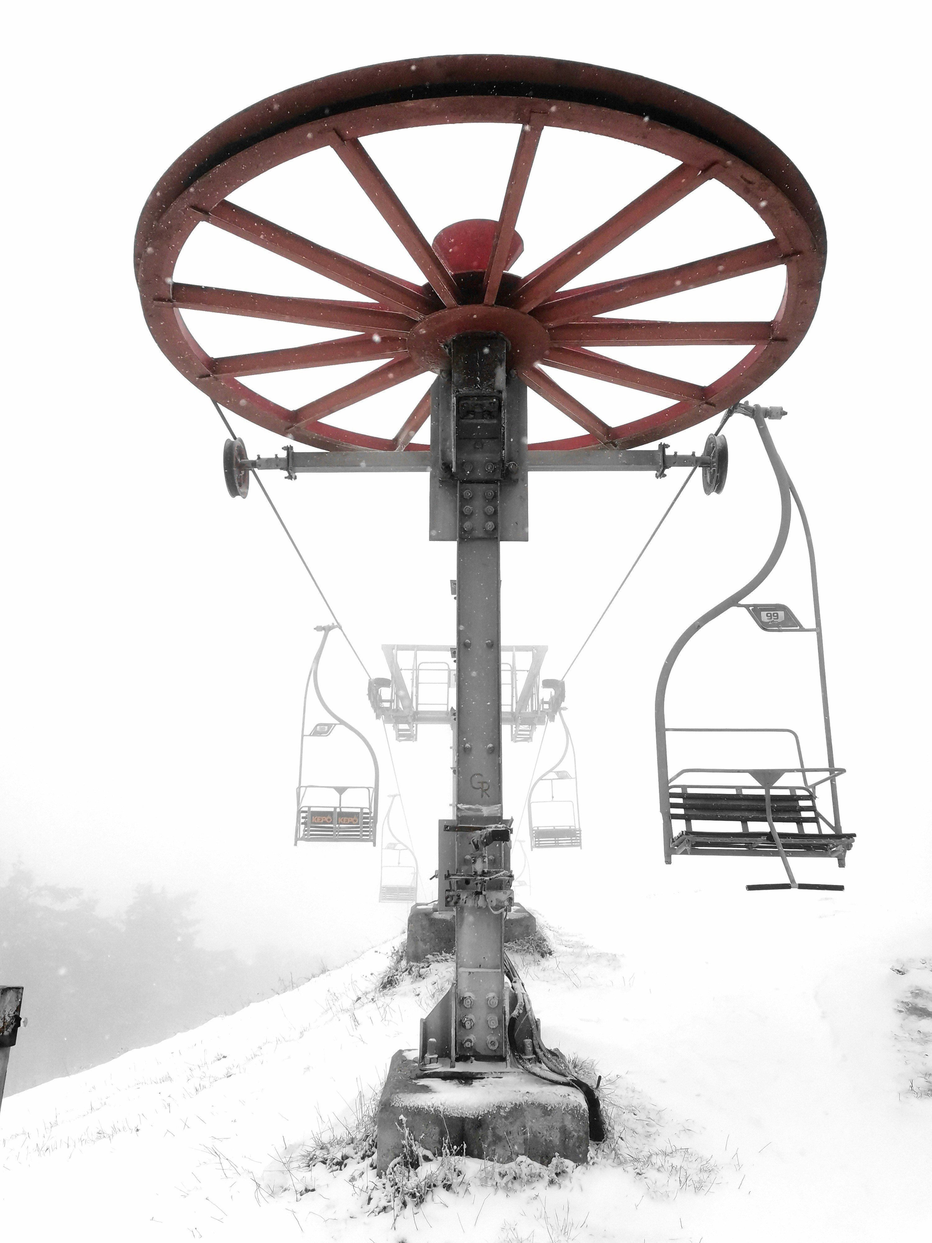 A snow-covered ski lift stands in a foggy landscape, its red wheel contrasting with the monochrome surroundings. The scene evokes a sense of solitude and anticipation.