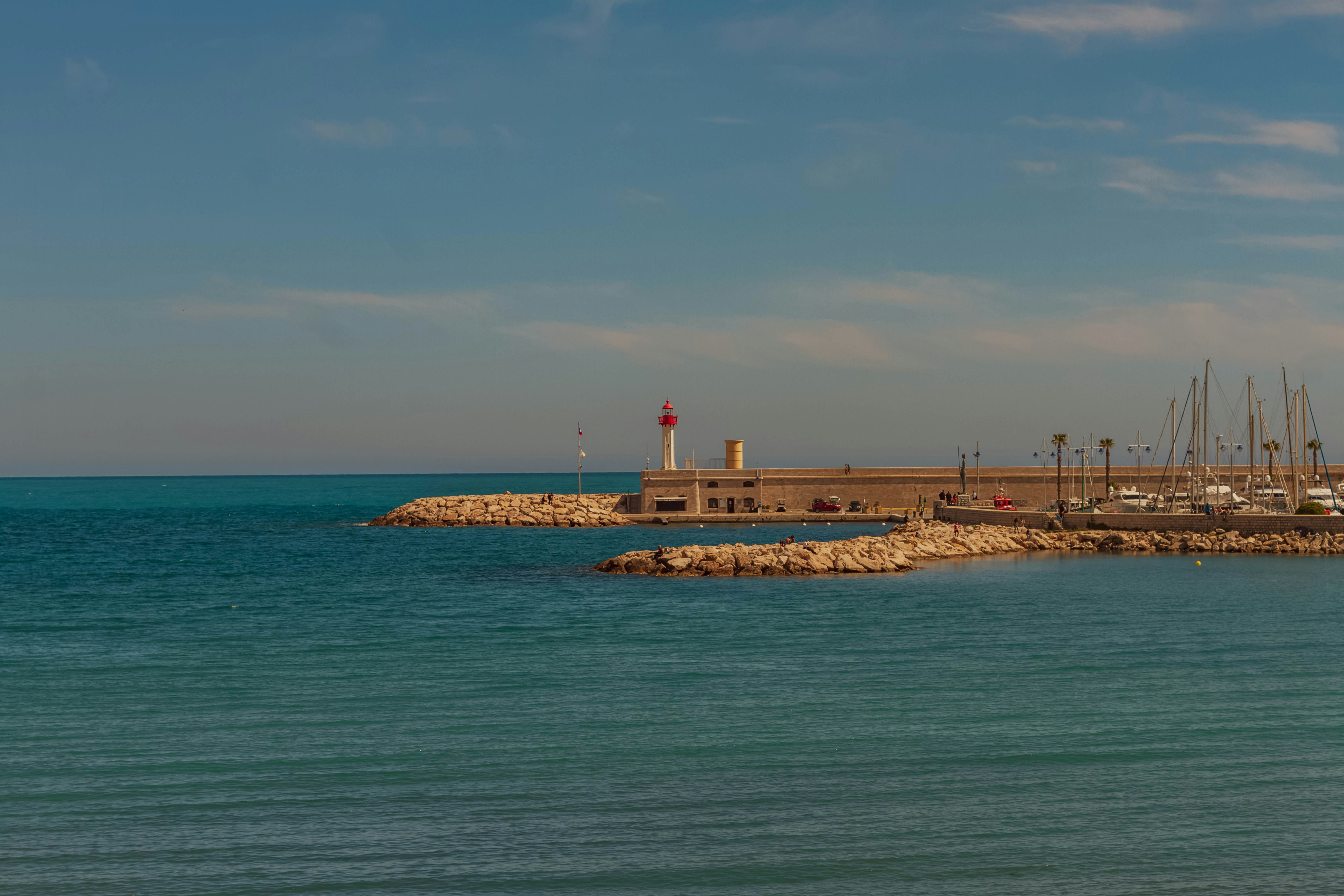 Lighthouse and distant sailboats along a serene Mediterranean coastline under a clear sky.