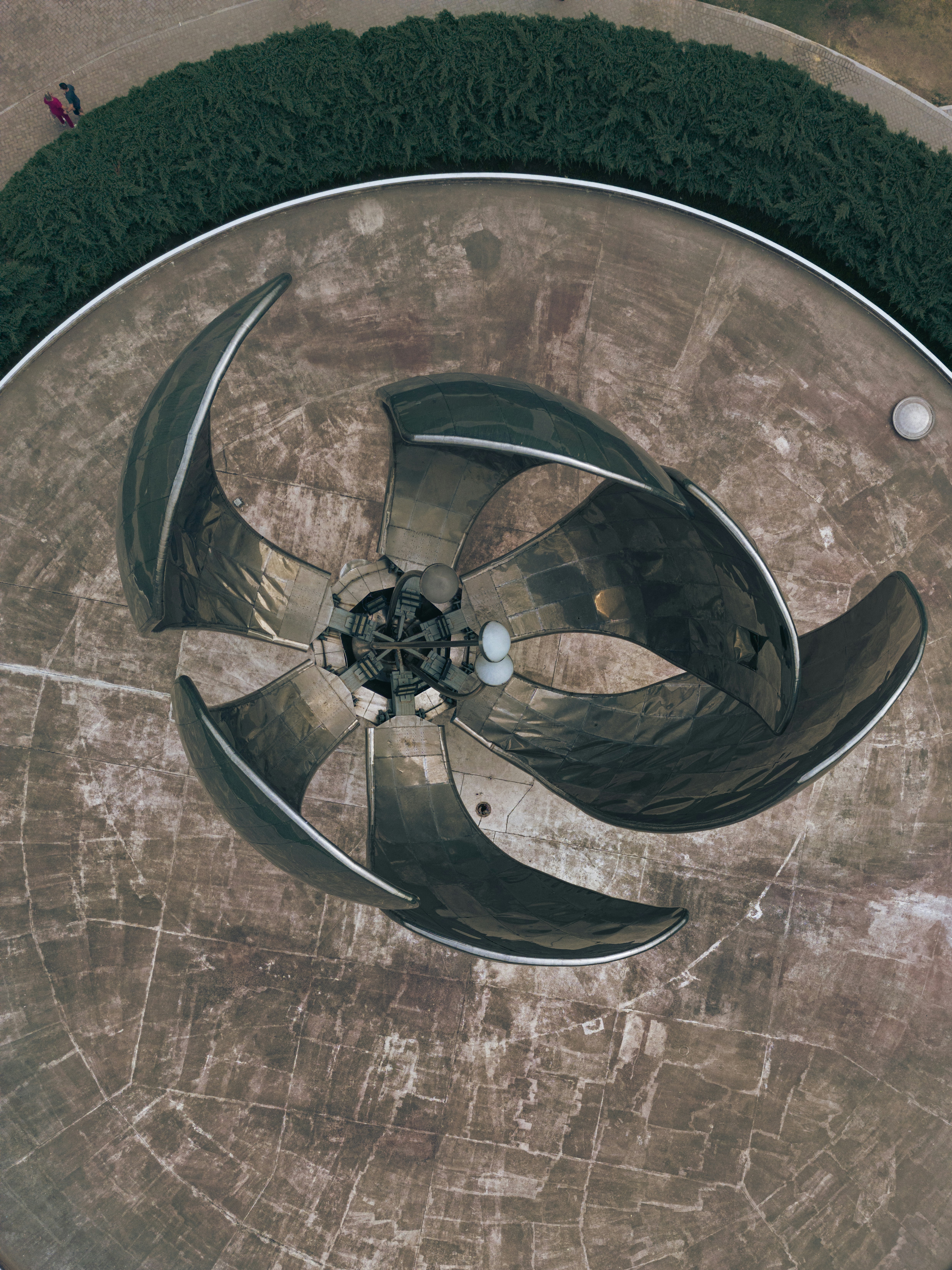 Top-down view of a metallic, spiral structure surrounded by a circular stone platform and greenery.