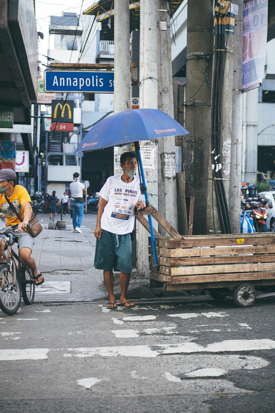 Man standing on a busy city street beside a wooden cart and blue umbrella, with power poles, wires, and a street sign overhead. A cyclist passes in the foreground amid the urban clutter.