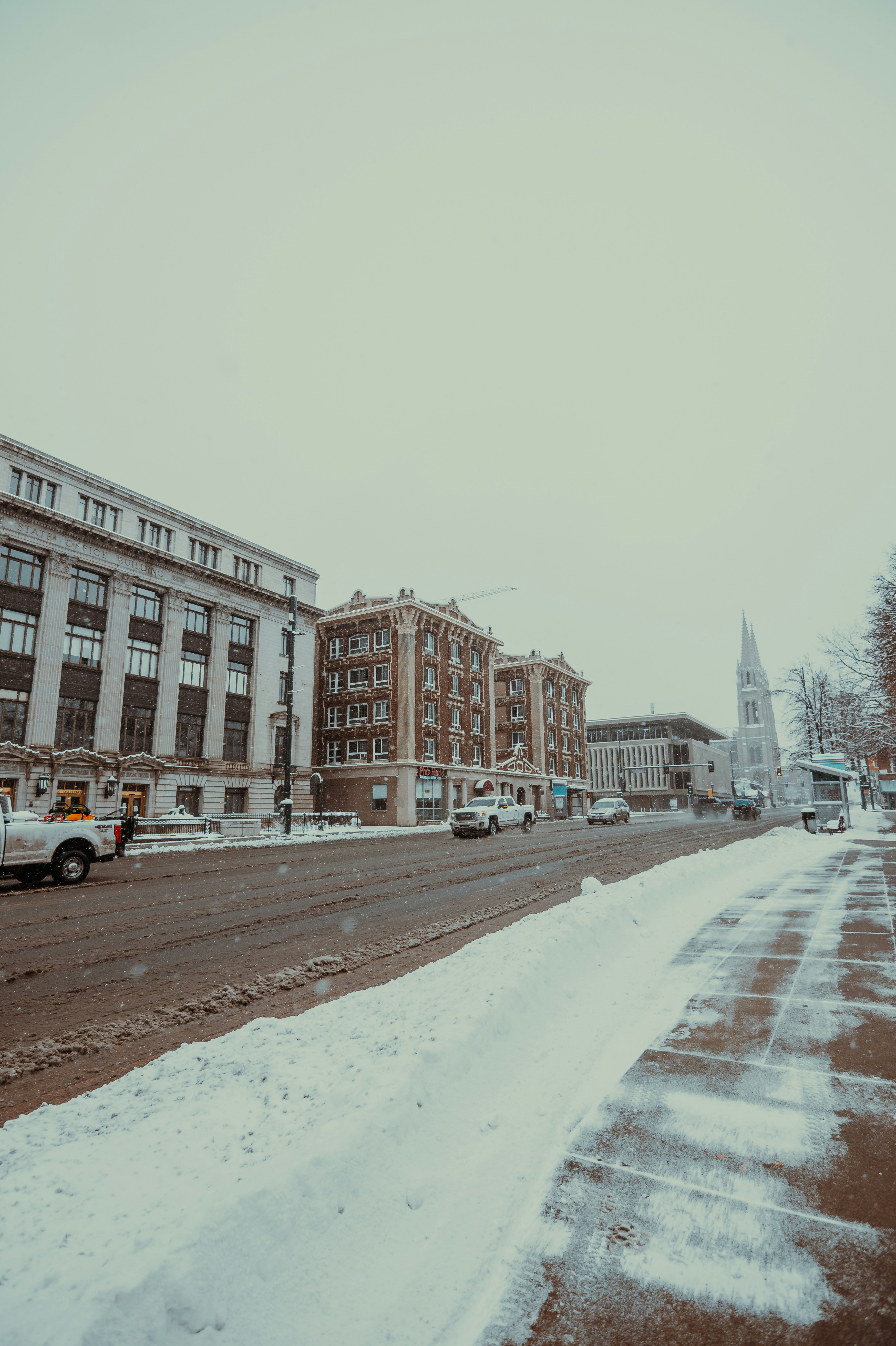 Snow blankets the streets of a quiet city, with historic buildings standing resilient against the winter chill. A distant church spire reaches towards the gray sky.