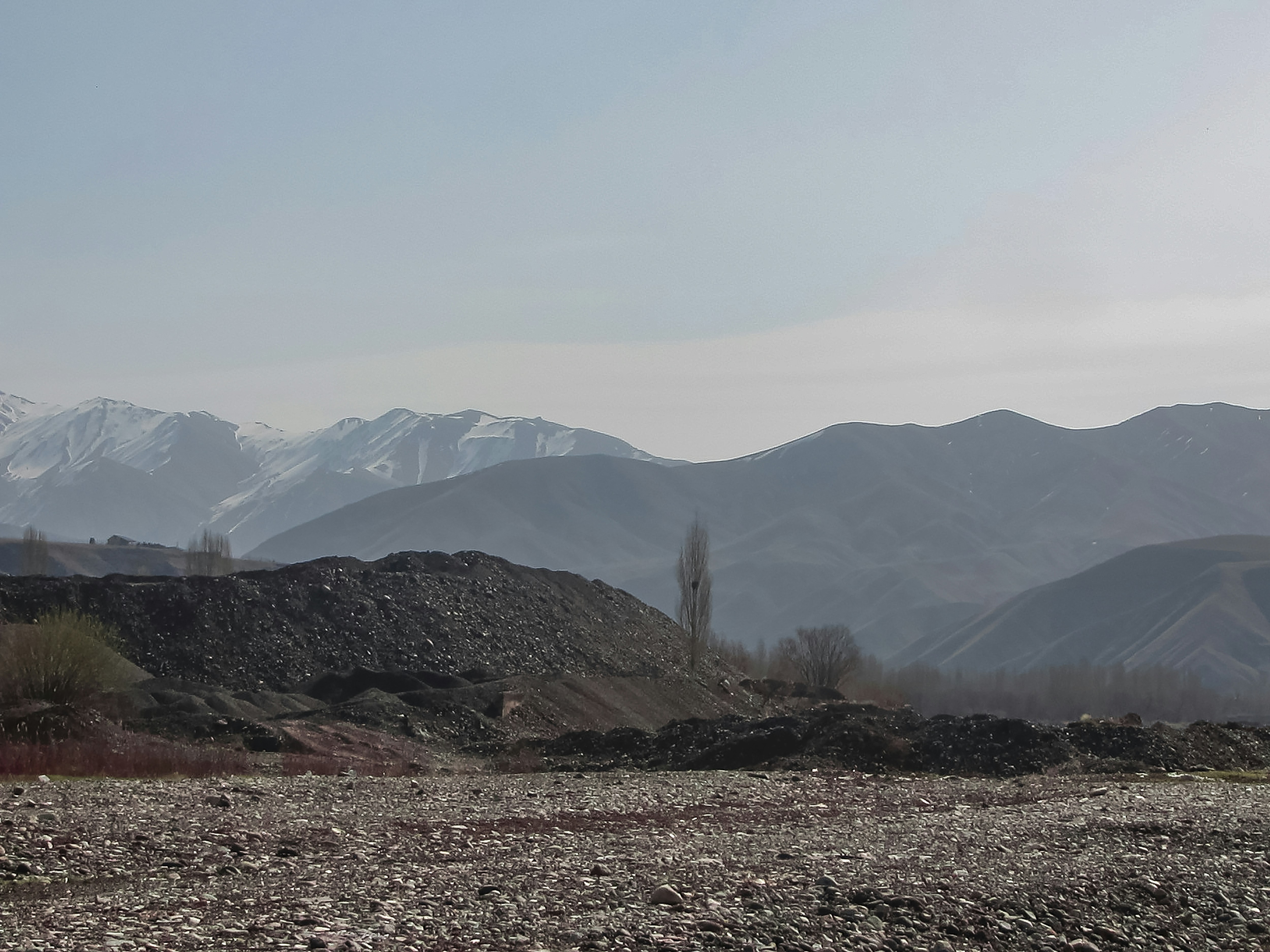 Wide landscape with a gravel foreground and dark mounds, leading to distant snow-covered mountains under a pale sky.