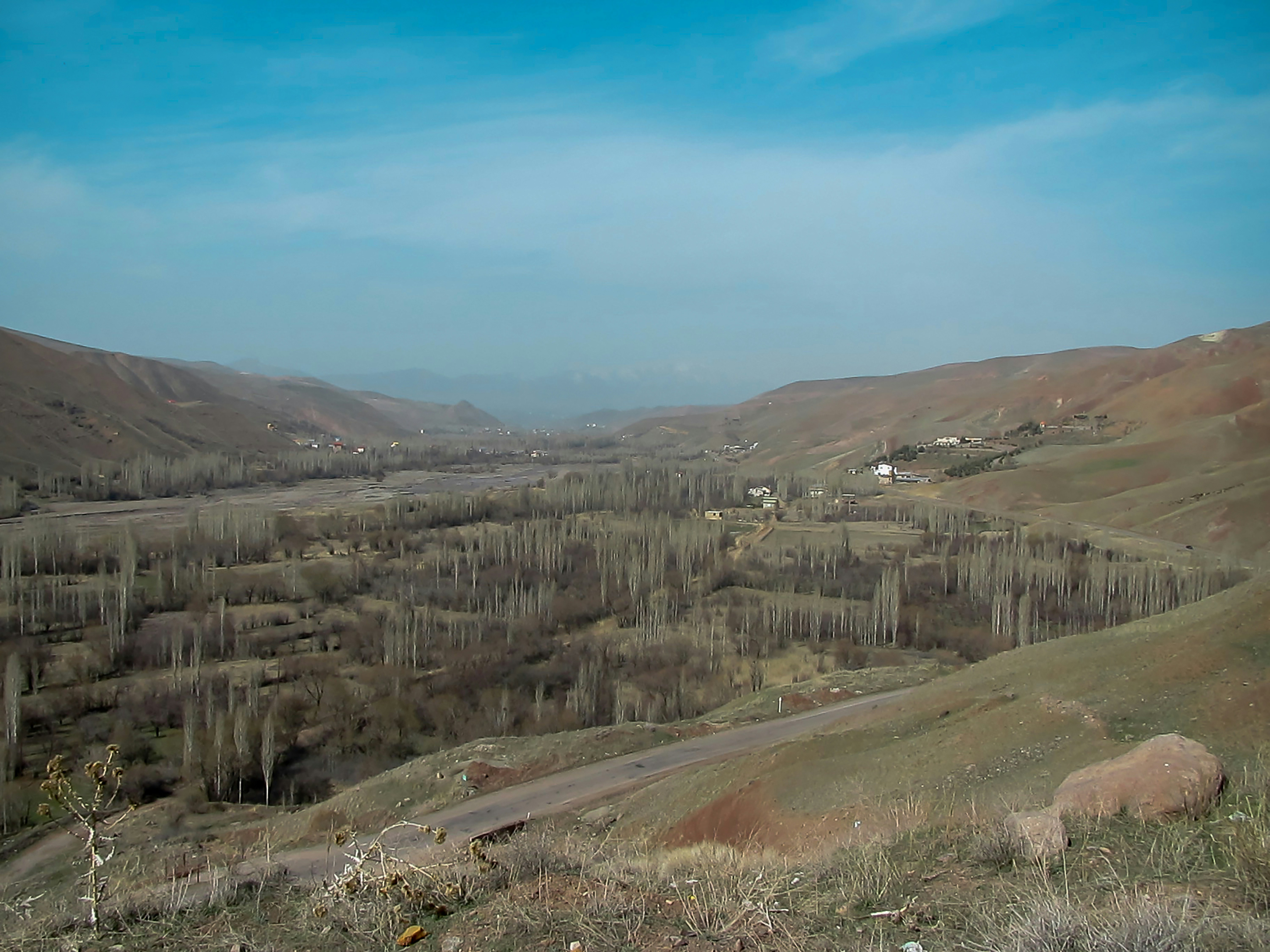 Wide valley scene with leafless trees along a winding road and distant houses, set among rolling brown hills under a pale blue sky.